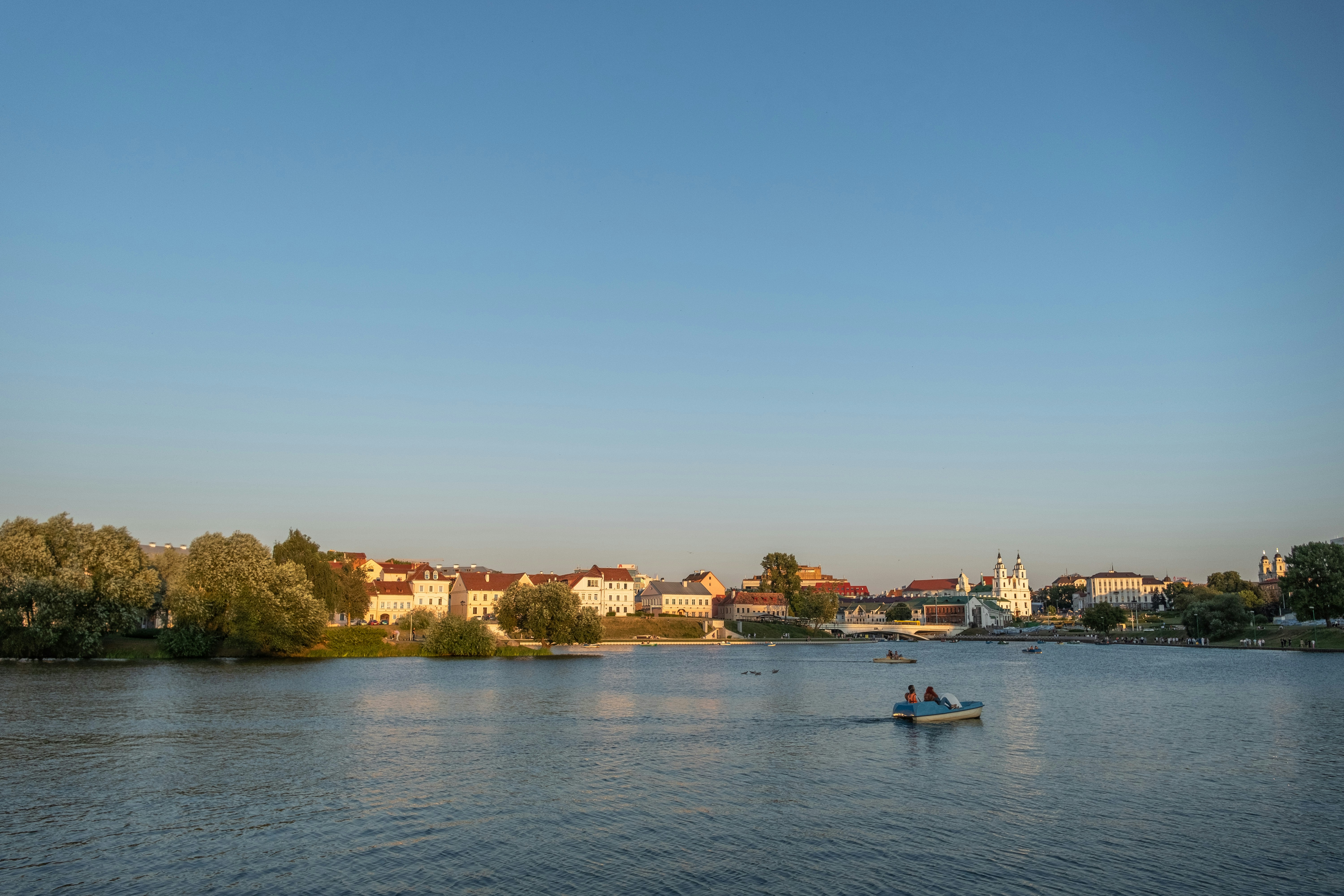 Small boat glides across a calm river against a distant town backdrop under a clear blue sky.