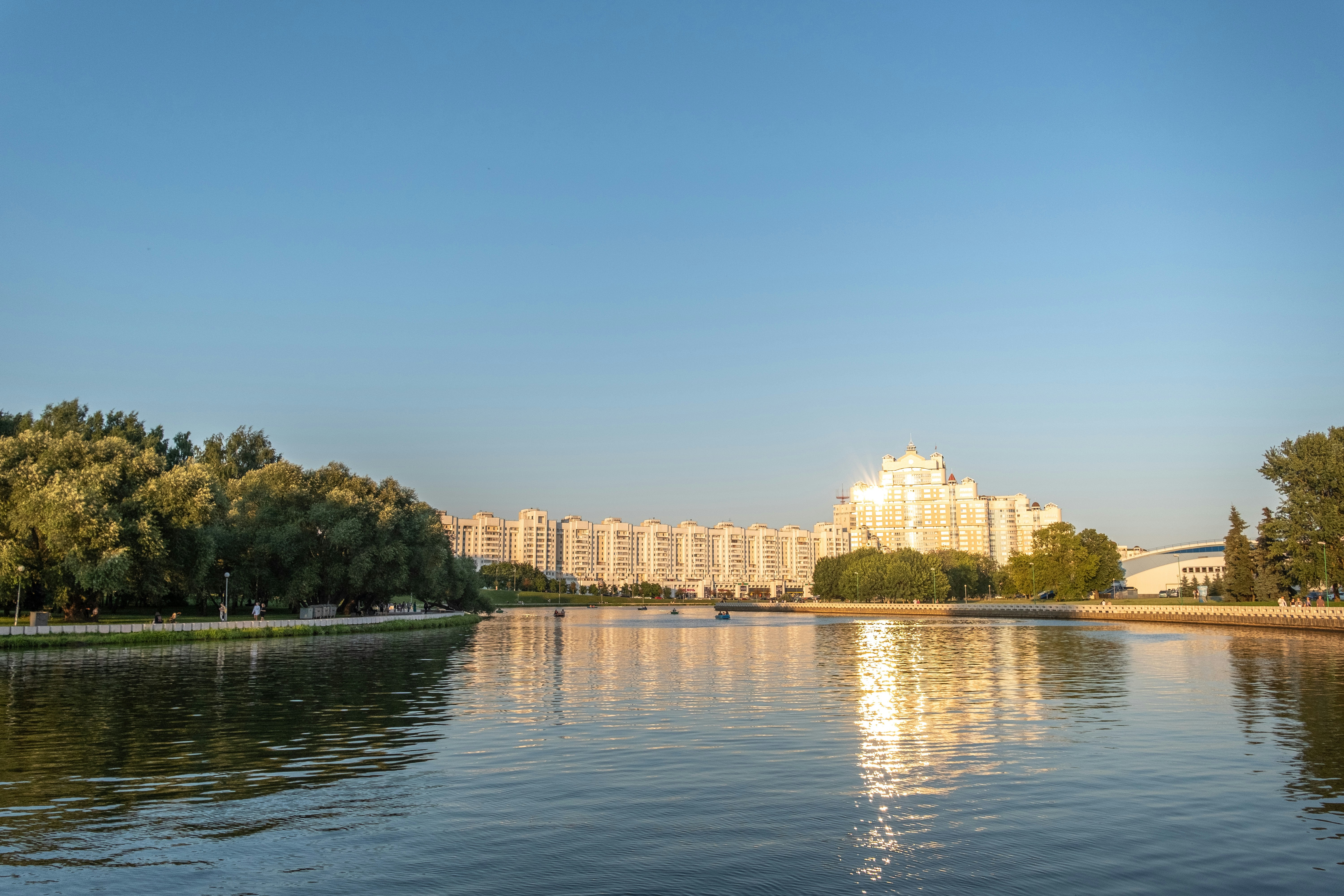 Sunlit buildings reflecting on a tranquil river, framed by lush greenery along the bank.
