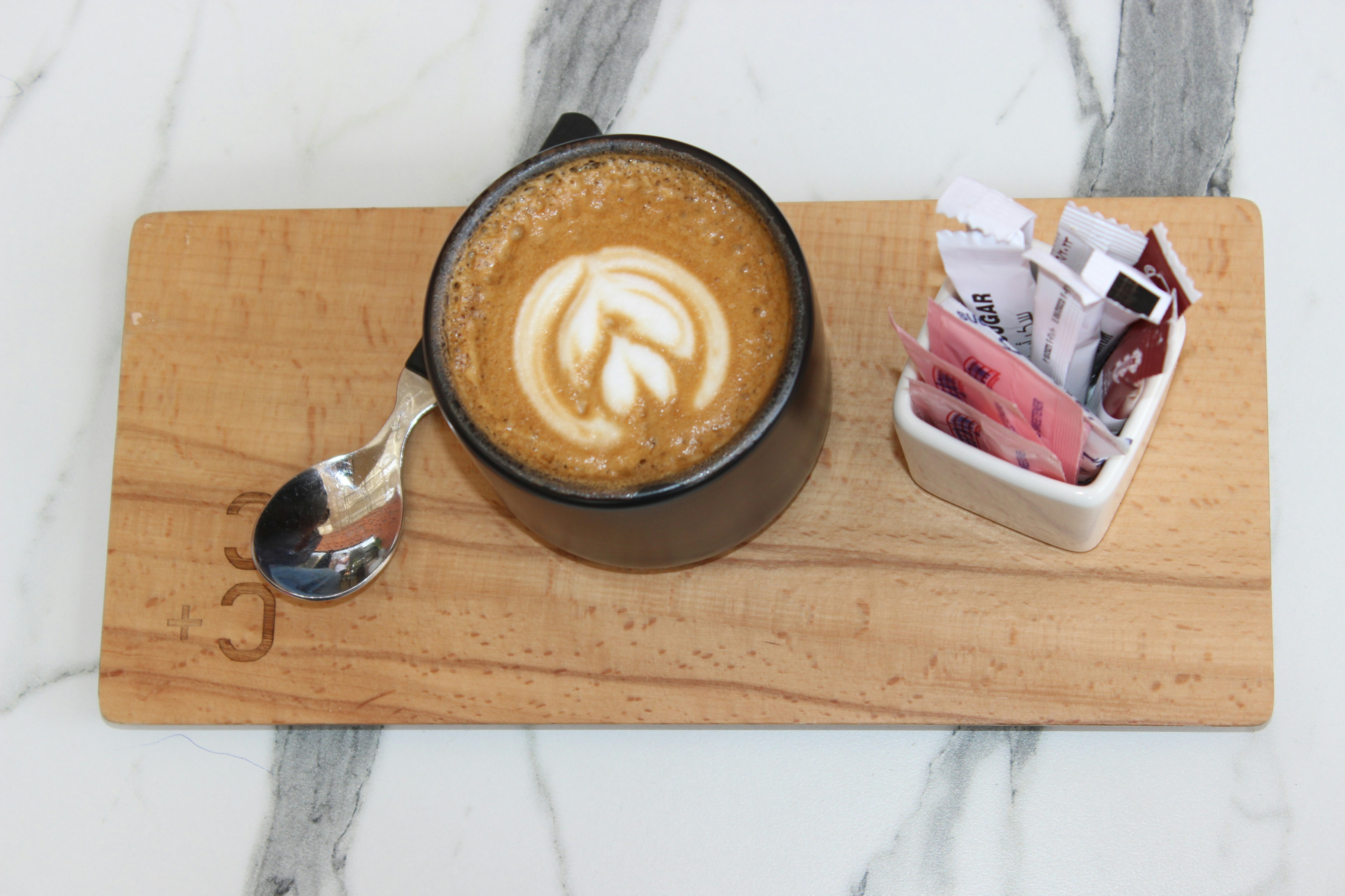 A beautifully crafted cup of coffee with intricate latte art sits on a wooden board, accompanied by a selection of sugar packets in a small container.