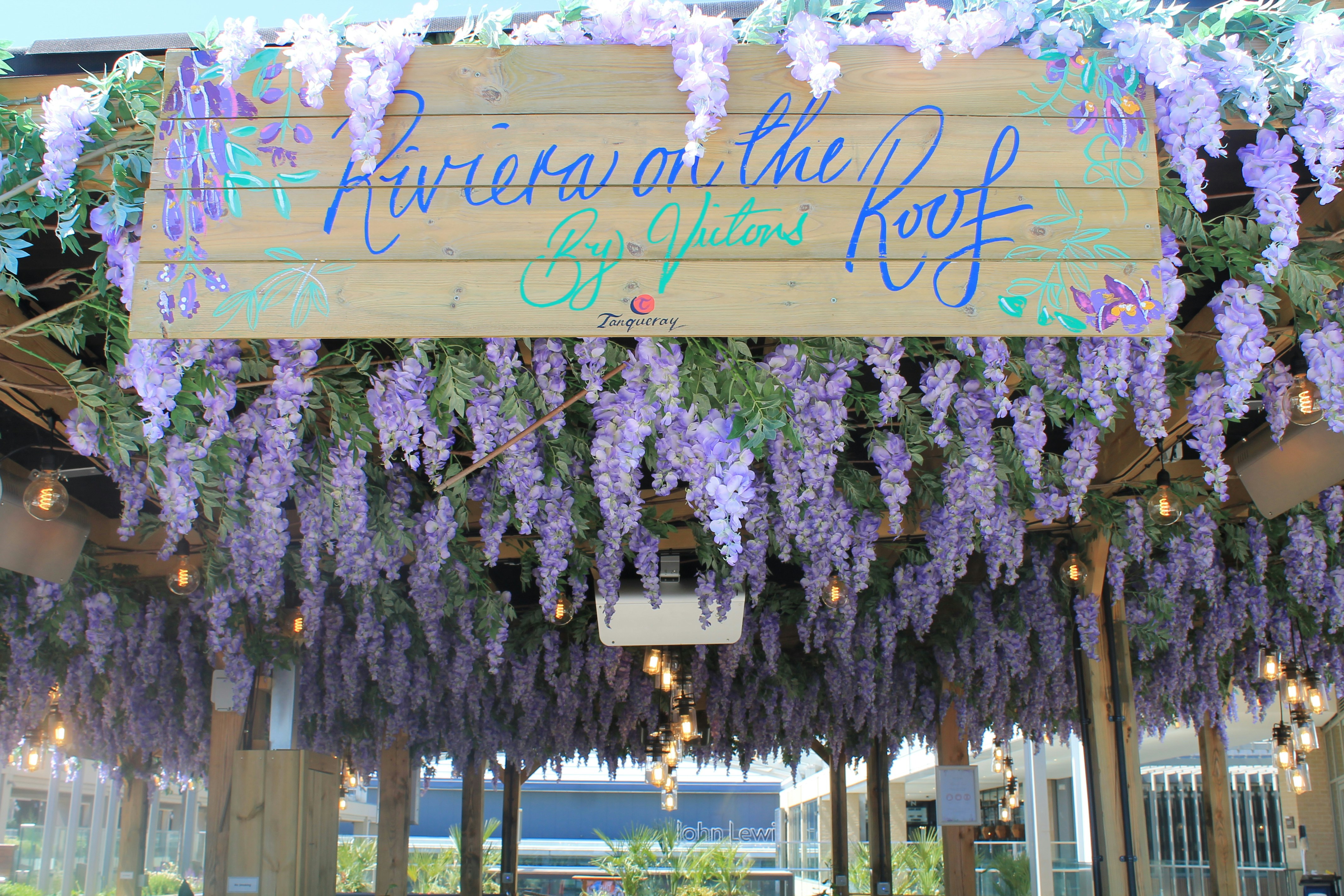 Vibrant purple wisteria drapes over a wooden sign that reads 'Riviera on the Roof,' inviting patrons to enjoy a unique rooftop experience.