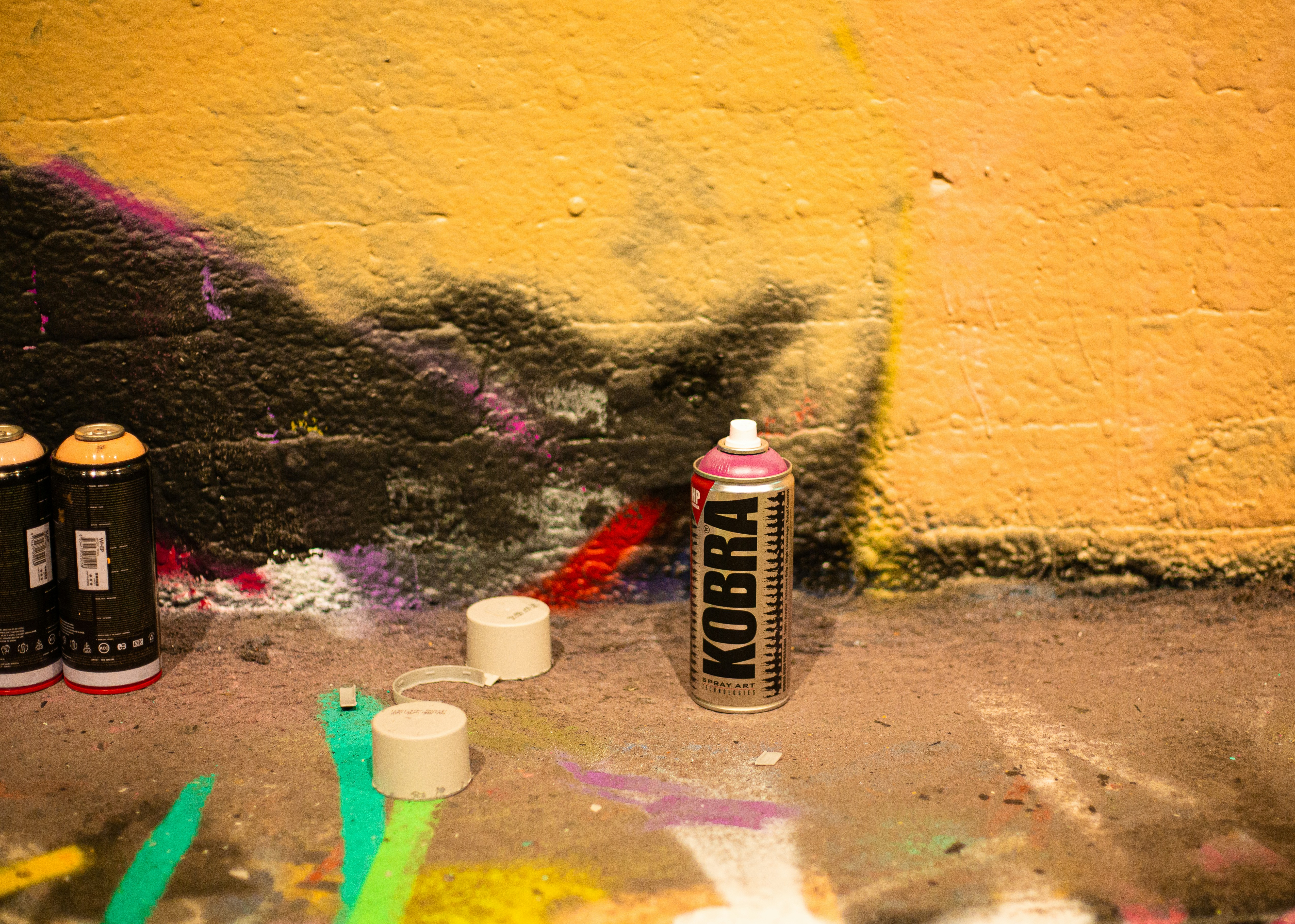 A spray paint can labeled 'KOBRA' stands prominently against a vibrant, graffiti-covered wall, surrounded by colorful paint splatters on the ground.