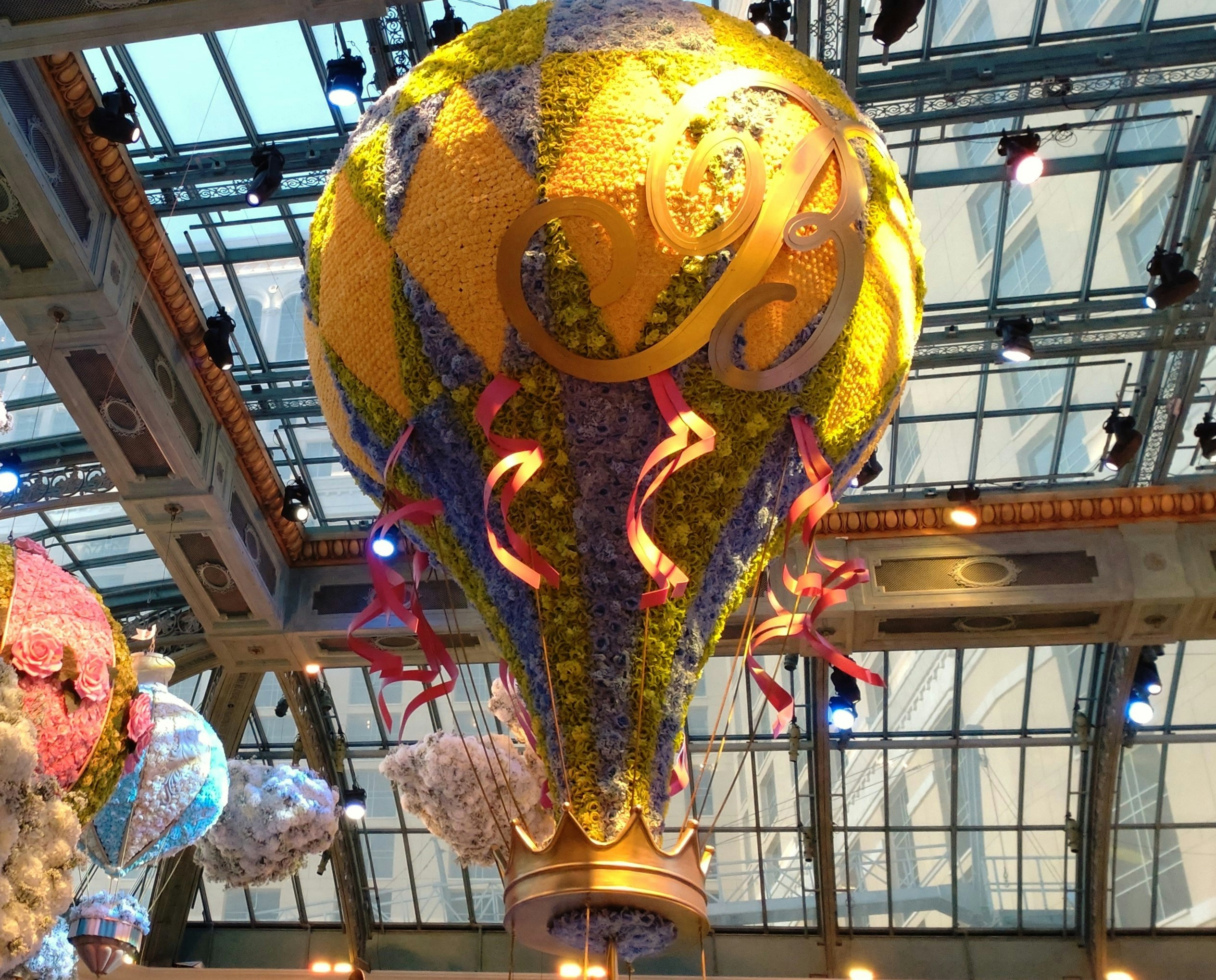 Colorful ornate balloon medallion suspended in a glass-ceiling atrium with ribbons and ambient lighting.