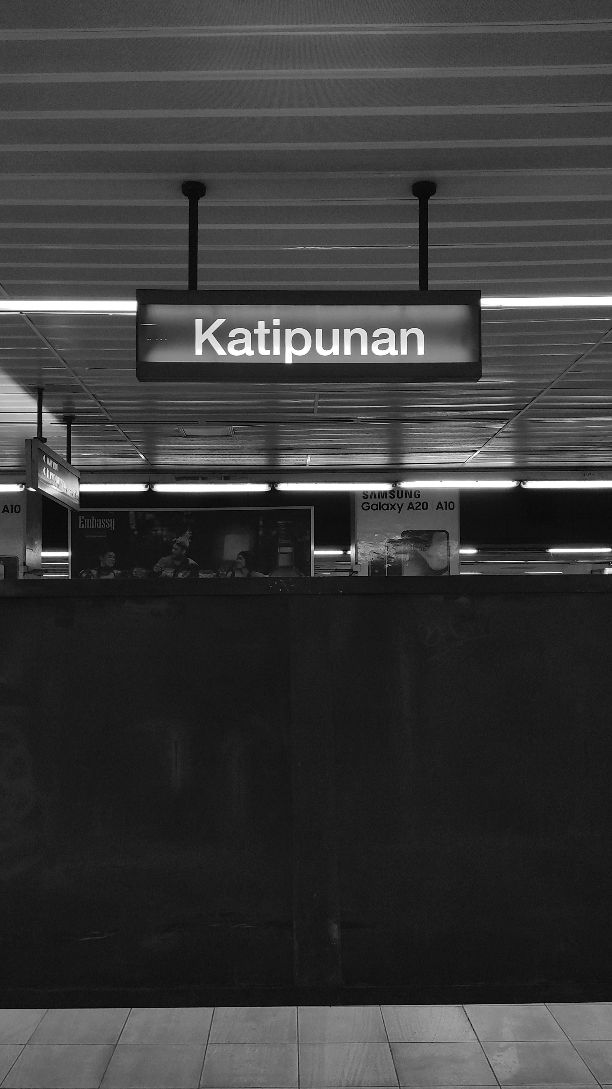 Black-and-white photograph of a Katipunan station sign suspended above a dark platform, with fluorescent lights and advertising boards in the background.