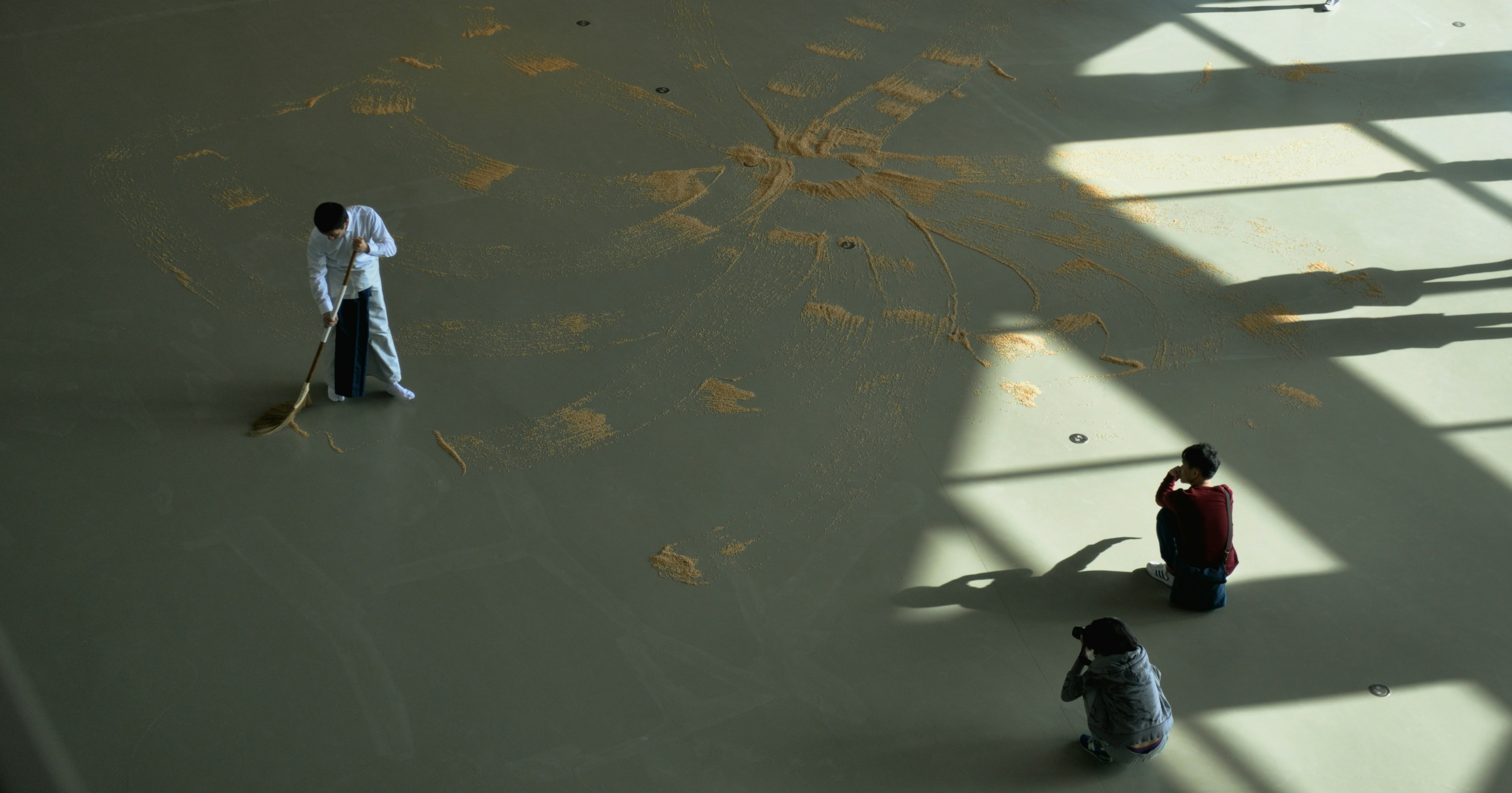 Overhead photograph of an indoor atrium showing a person sweeping on the left and two people seated on the right, with bold diagonal shadows across a pale floor.