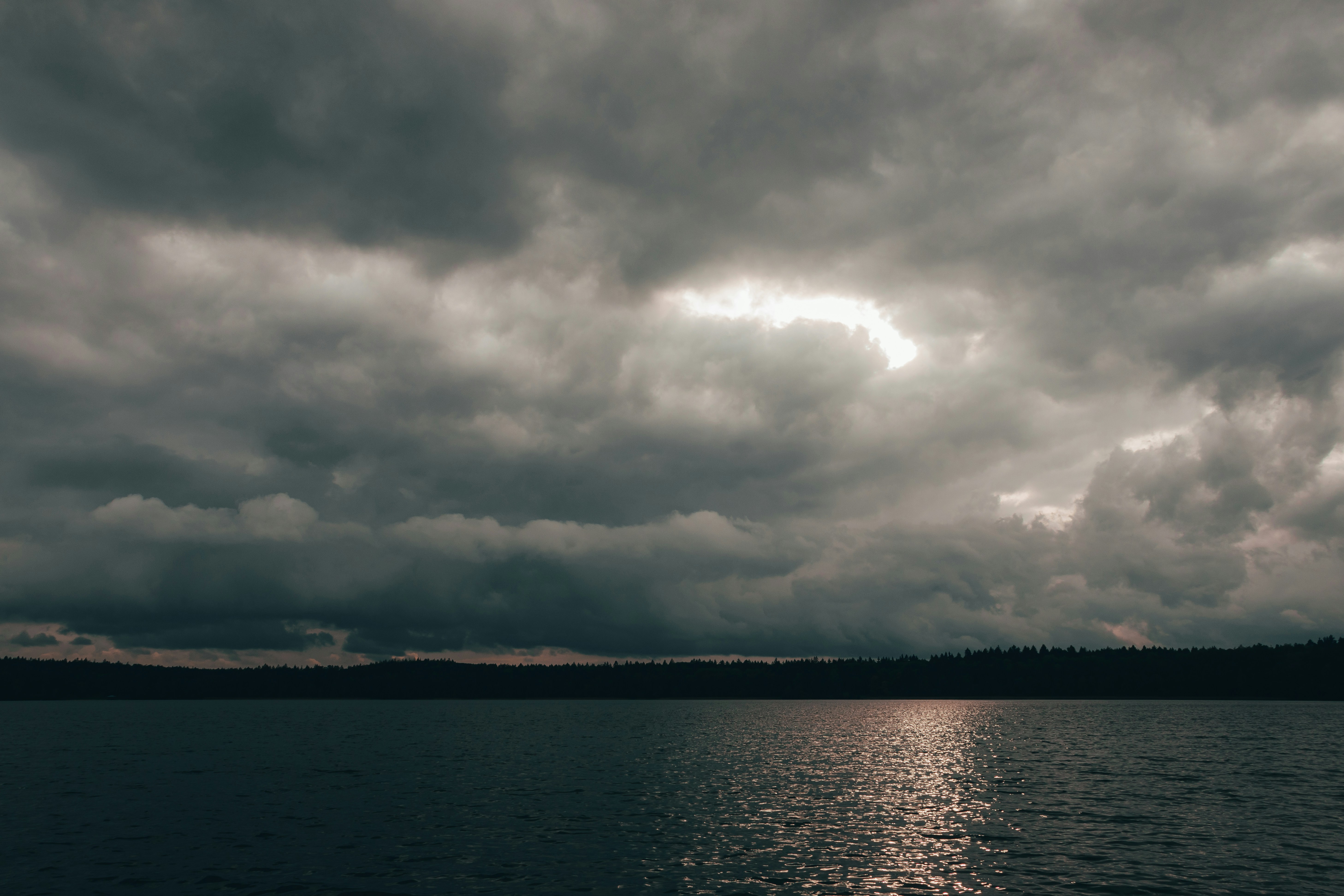 Dramatic clouds loom over a tranquil lake, reflecting the muted light of an overcast sky. The scene evokes a sense of calm before the storm.
