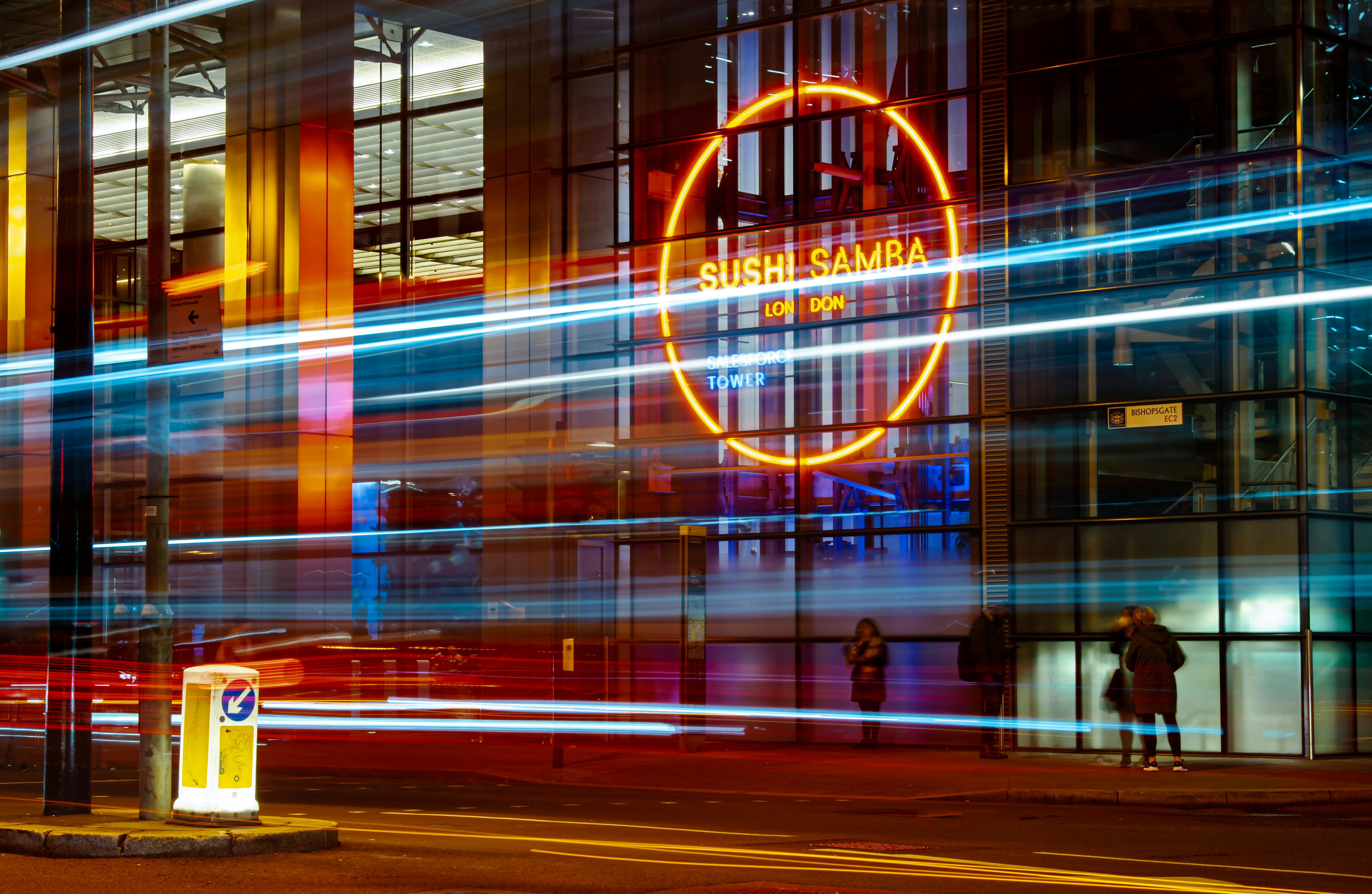 Light trails streak past a neon-lit building facade in an urban night scene.