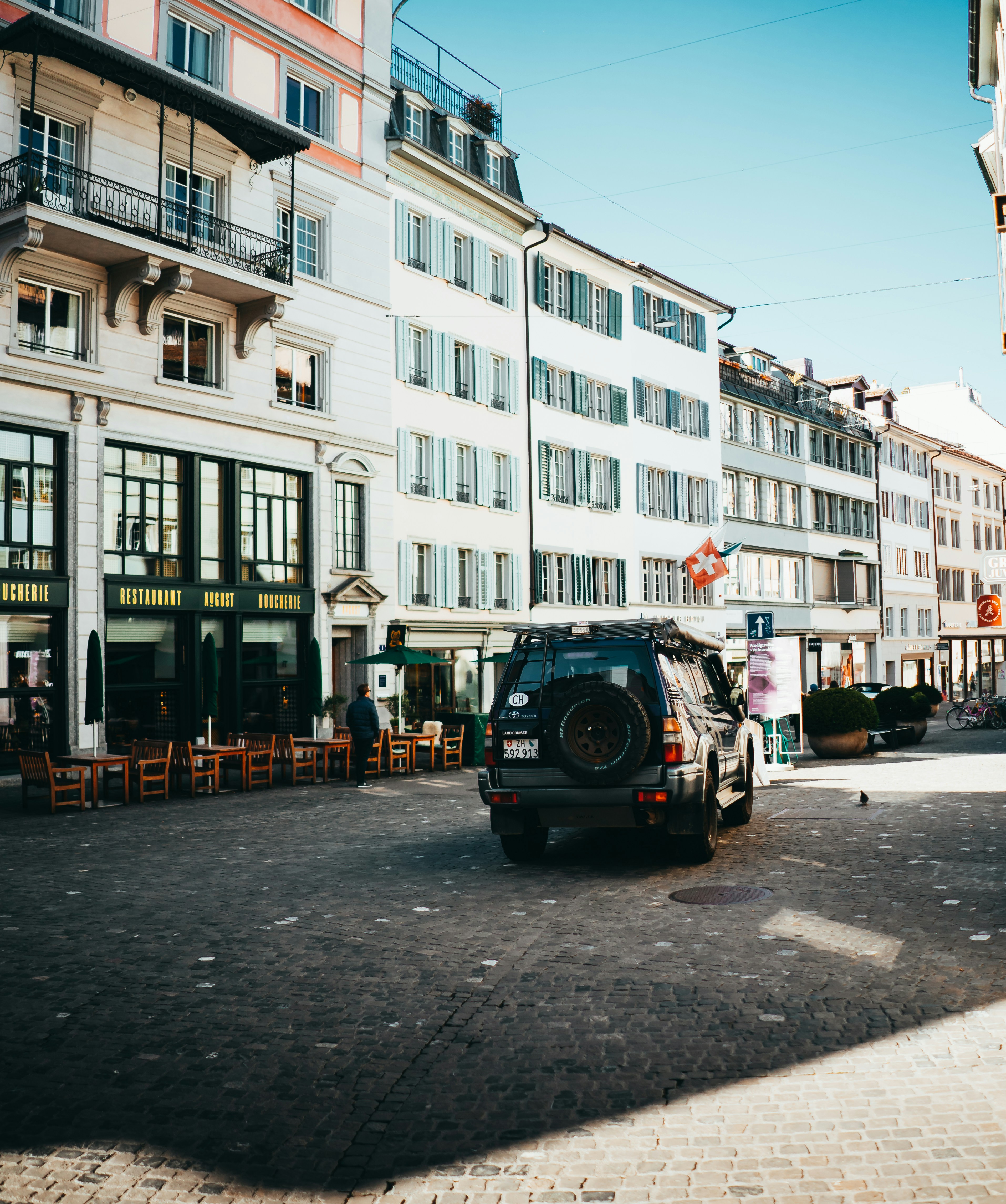 A parked vehicle on a cobblestone street, surrounded by charming buildings and outdoor seating, capturing a moment of urban tranquility.