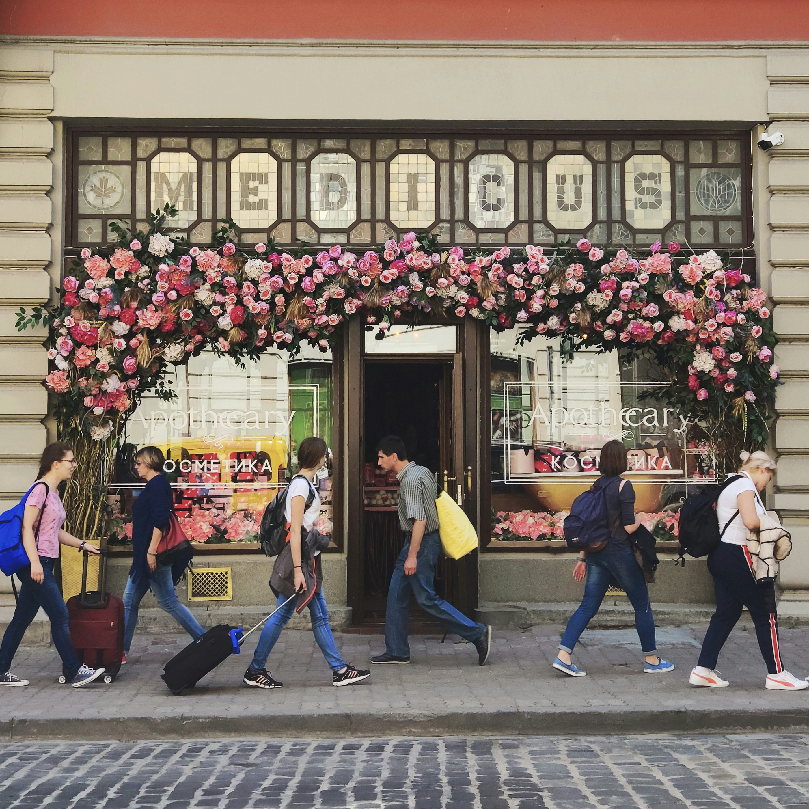 Bloom-Arch Crossing: Pedestrians Under a Flower-Framed Storefront