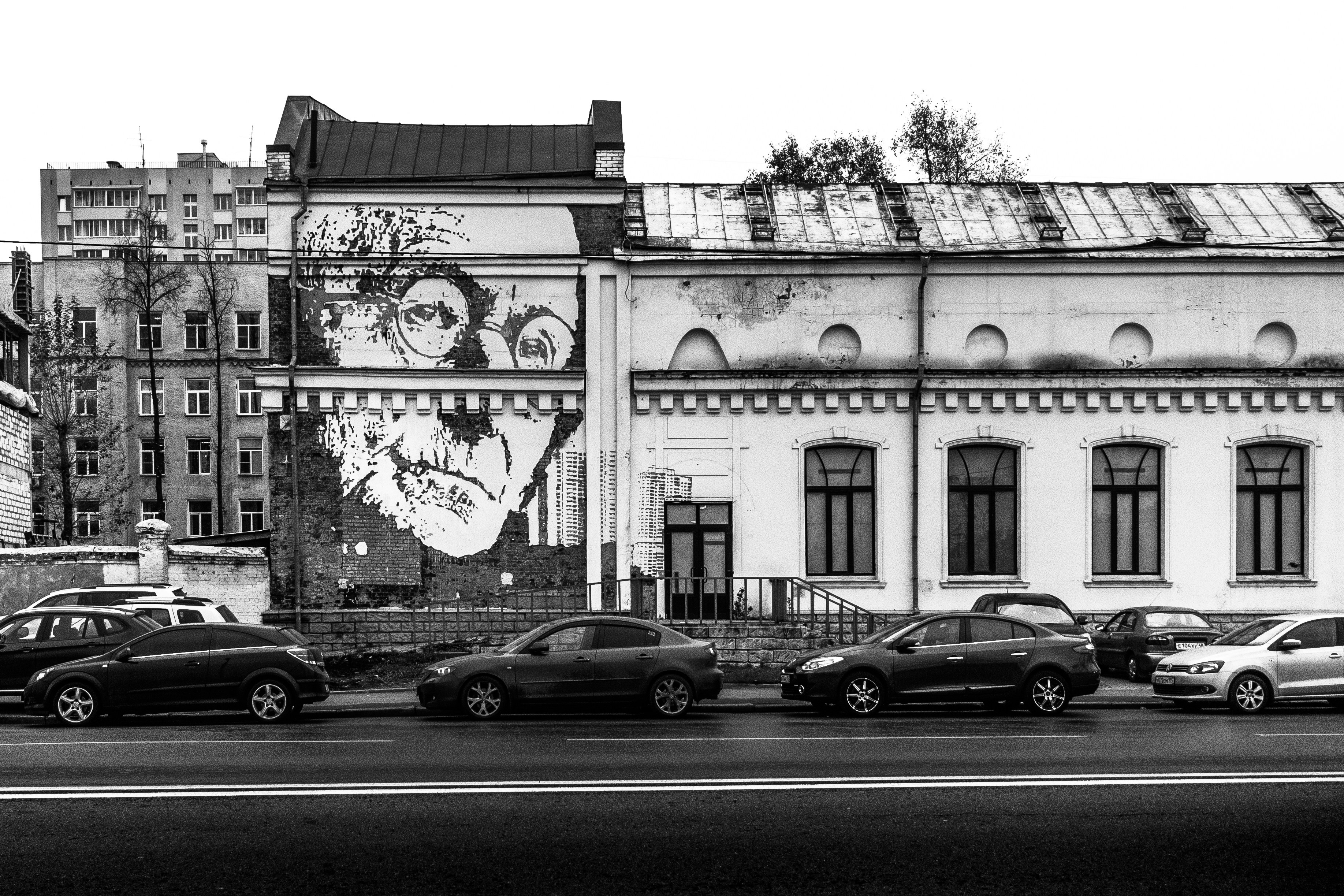 A striking mural of a face on a weathered building, juxtaposed with parked cars along the street. The scene captures the essence of urban art and architecture.