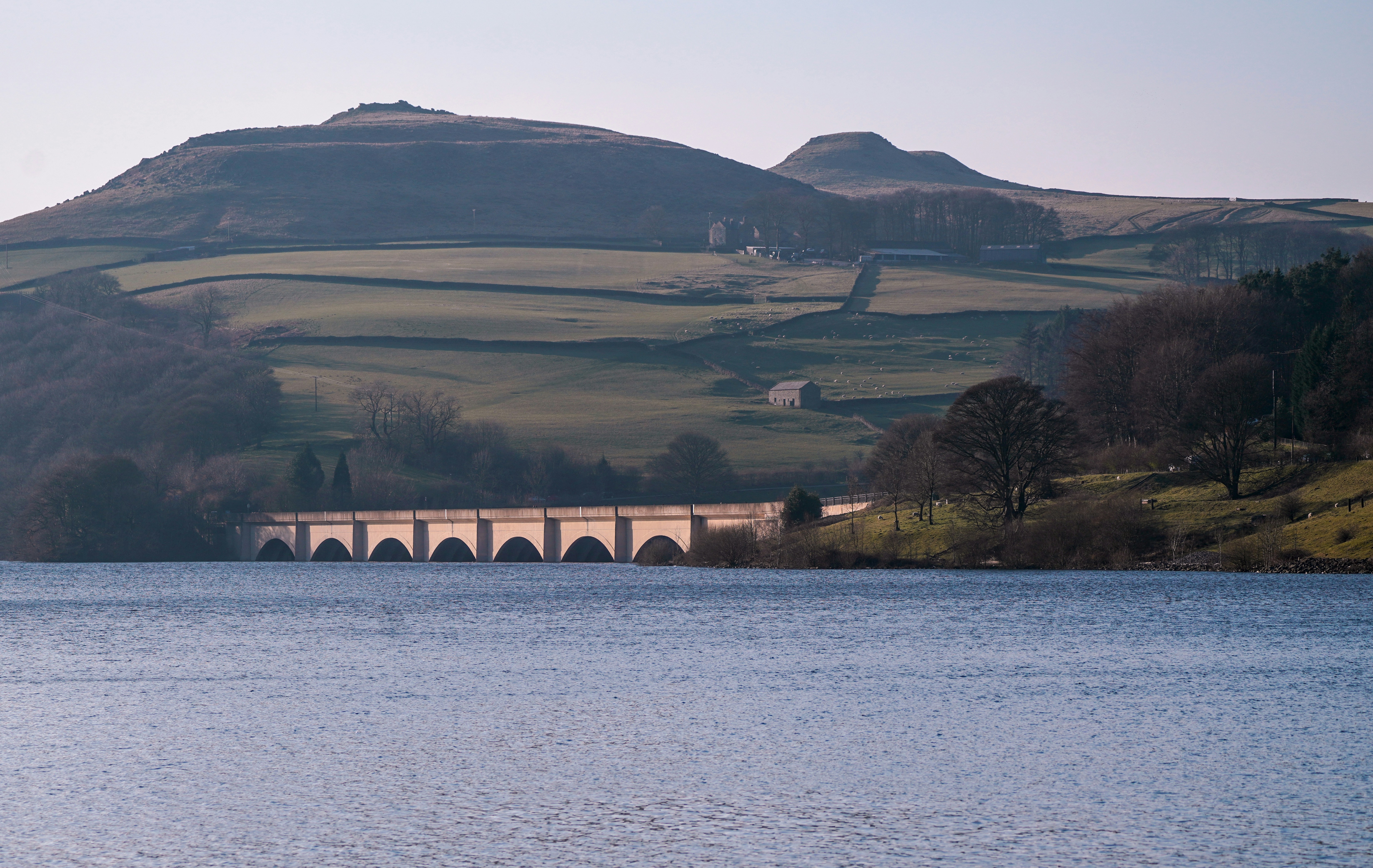 Historic stone bridge spanning a tranquil lake, framed by rolling hills and a clear sky.