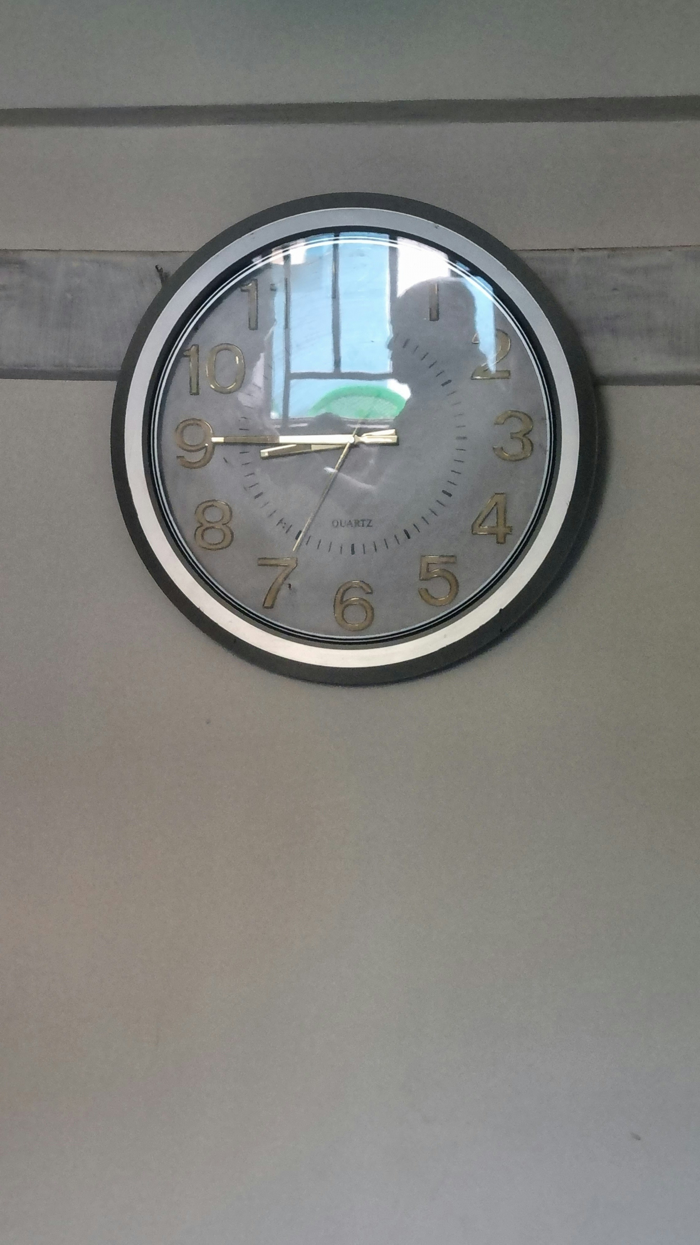 Wall clock displaying time with reflections of a window and greenery. The clock features a classic design with gold numbers against a muted background.
