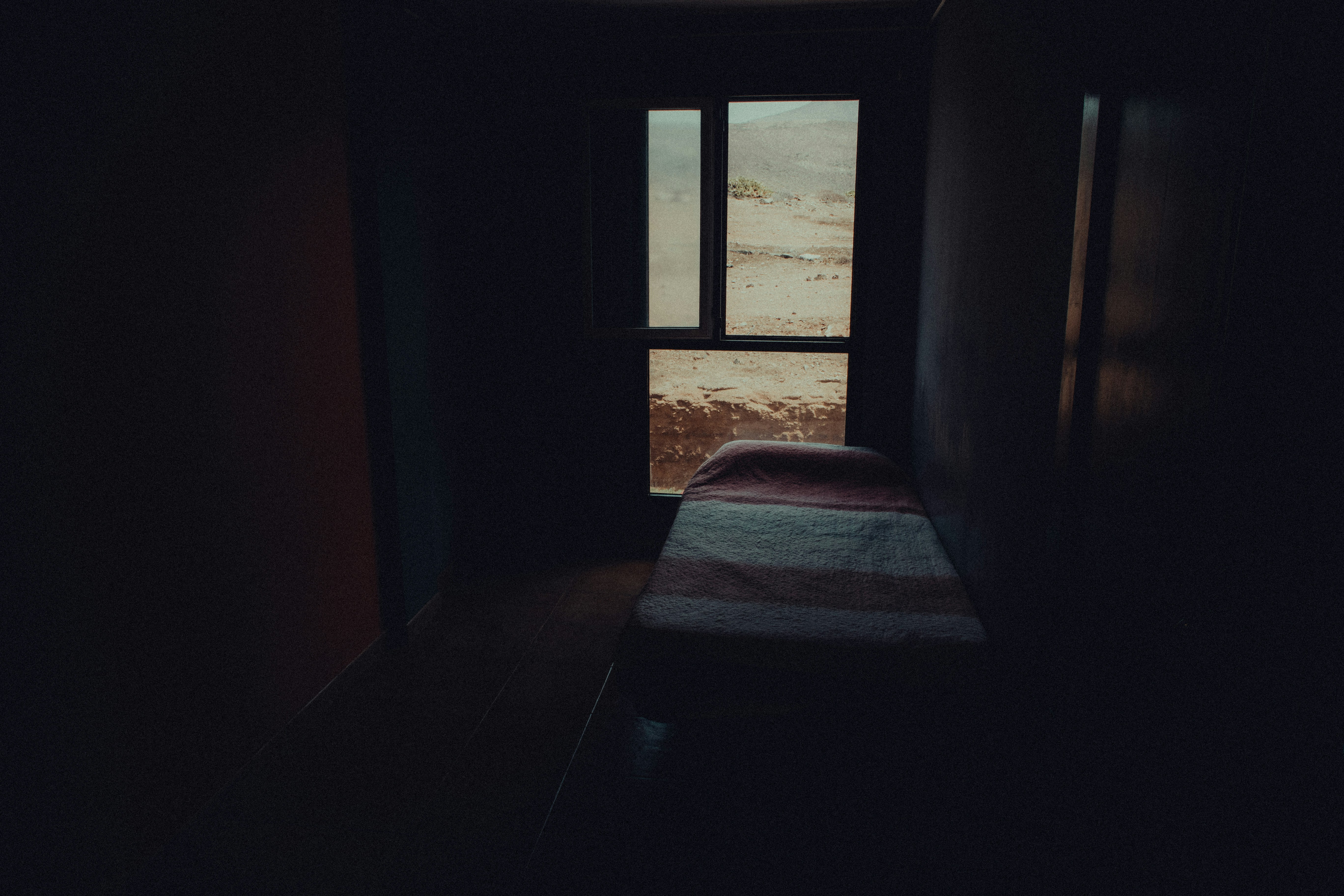 A dimly lit room featuring a bed and a large window that frames a distant landscape. The interplay of shadows and light creates a serene atmosphere.
