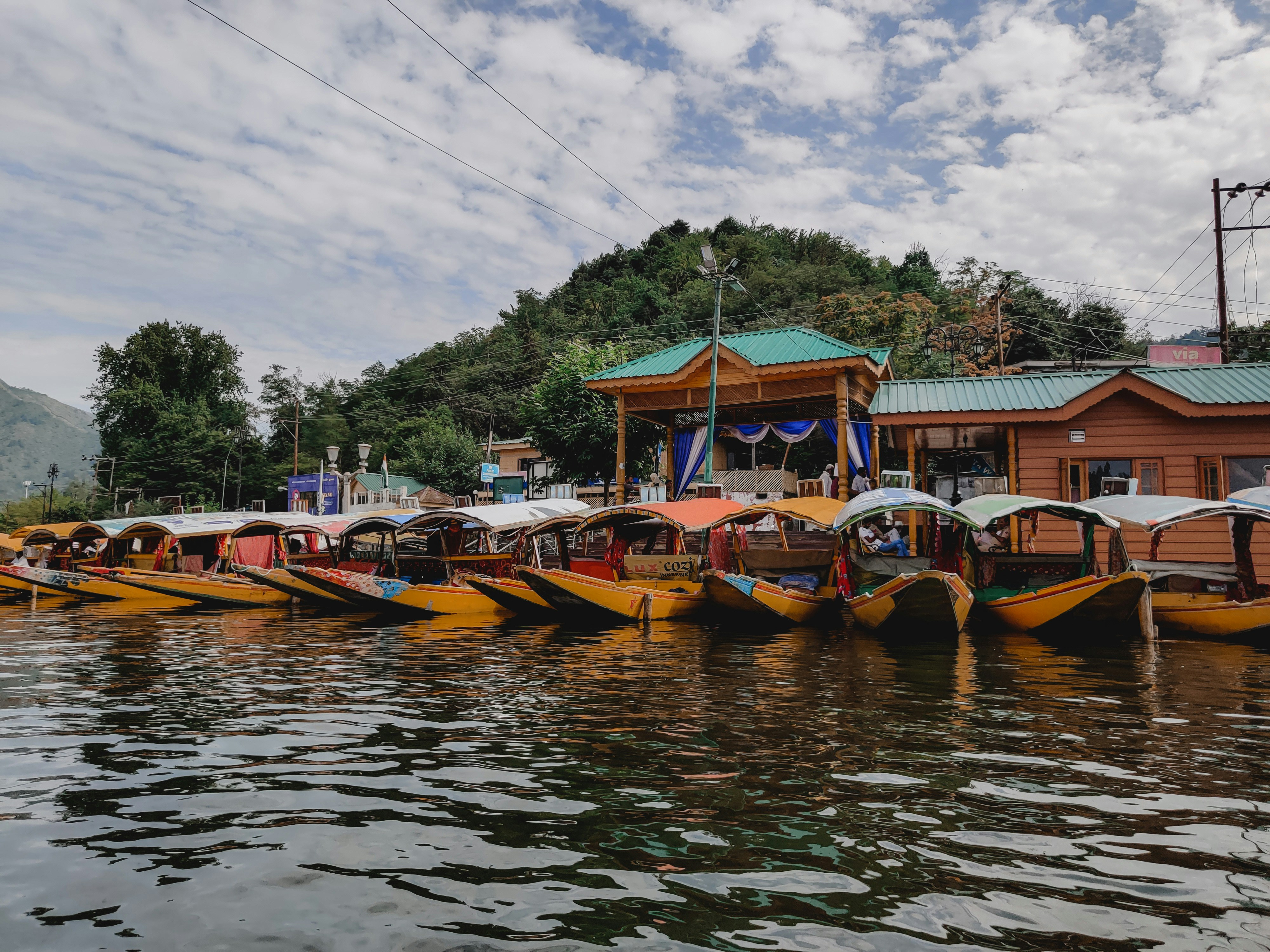 Row of colorful shikaras lined up on a serene lake with a backdrop of lush green hills under a partly cloudy sky.