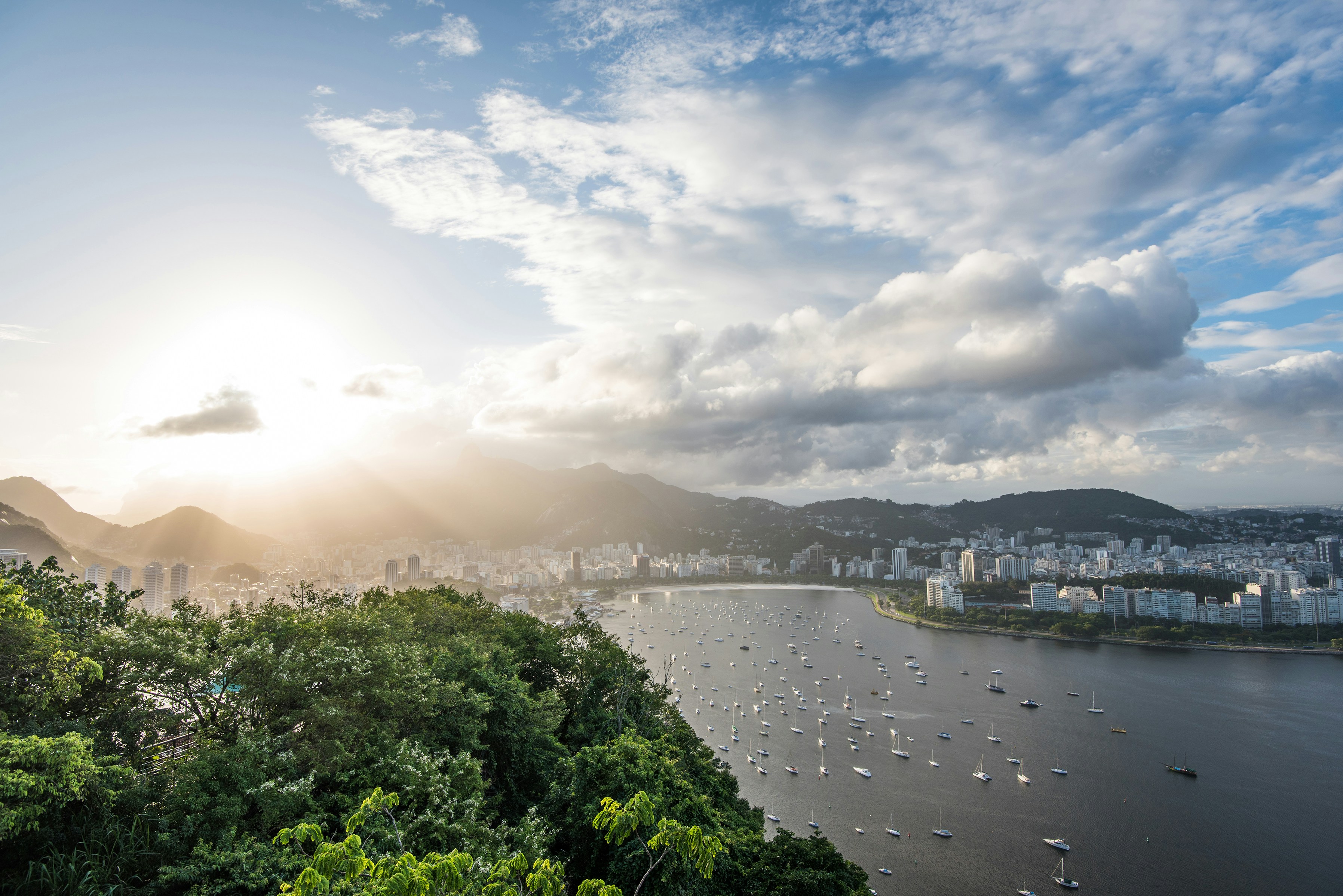 Panoramic view of a vibrant bay at sunset, with boats scattered across the water and a city skyline nestled against lush mountains.