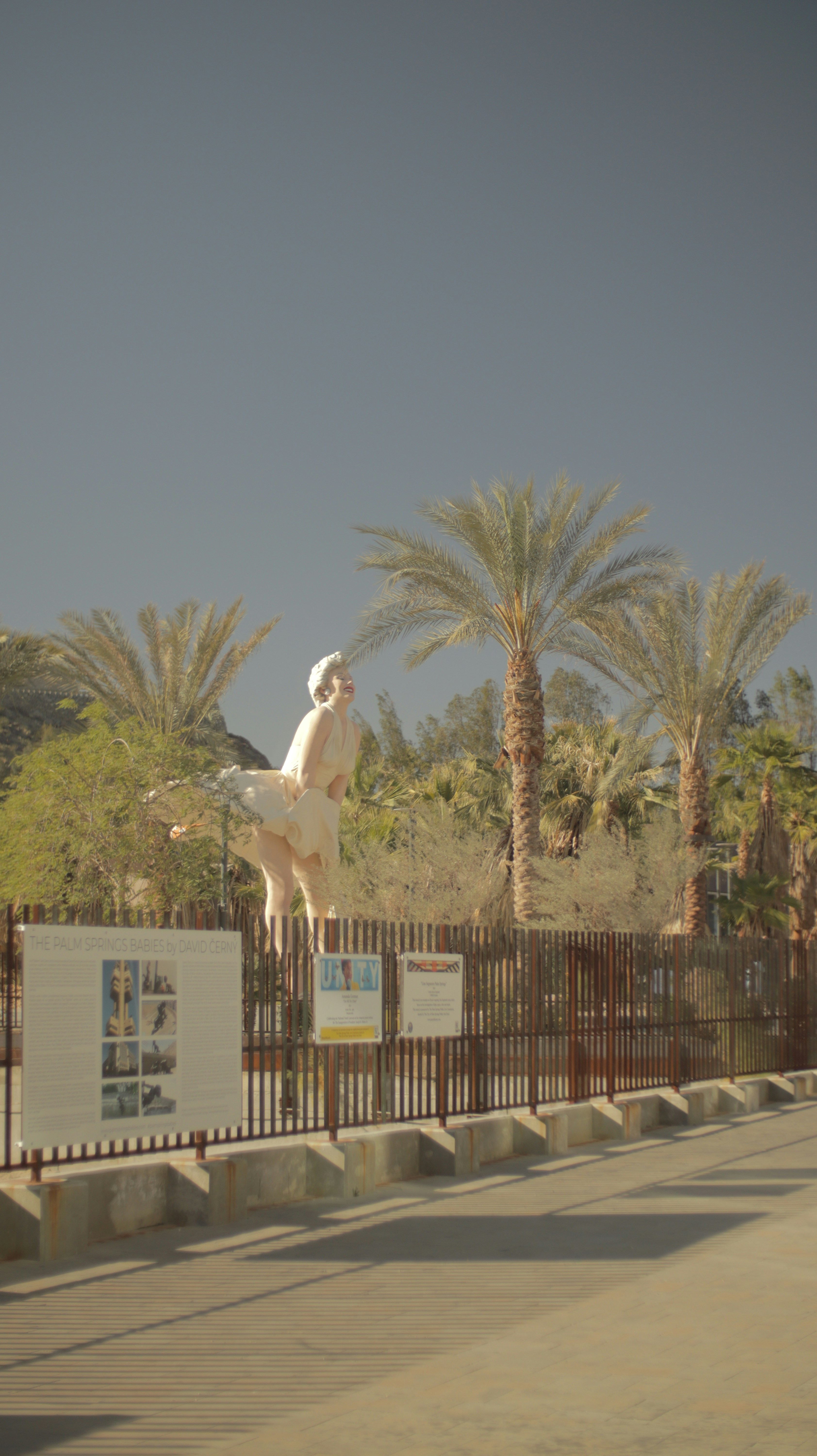 A large, playful statue stands amidst palm trees in a sunlit desert landscape, embodying a quirky charm. Informational signs are visible nearby.