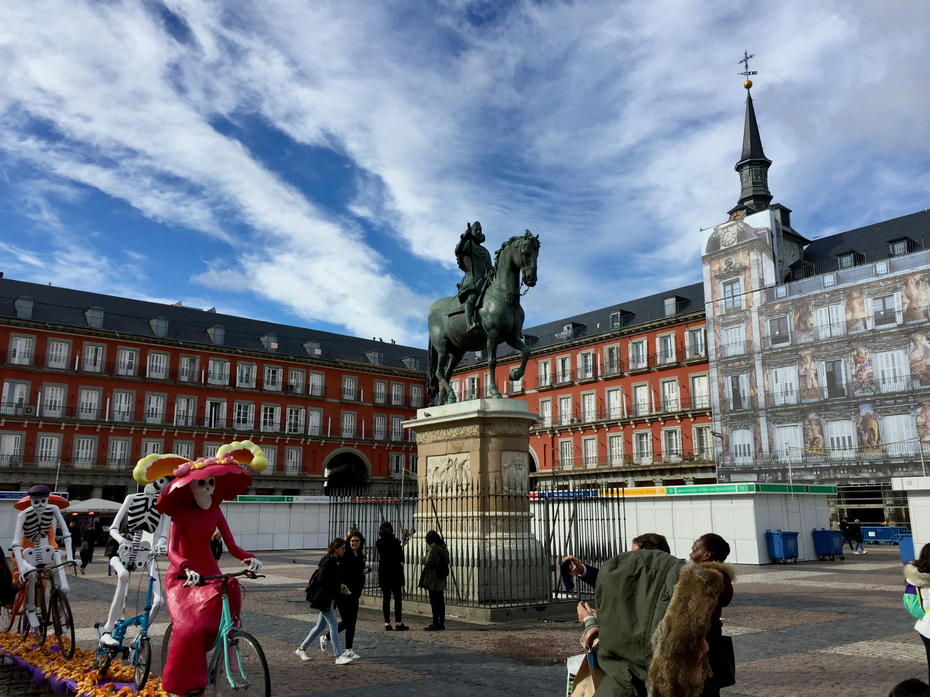 Colorful skeletons on bicycles celebrate Día de Muertos beside a historic statue in Madrid's Plaza Mayor.