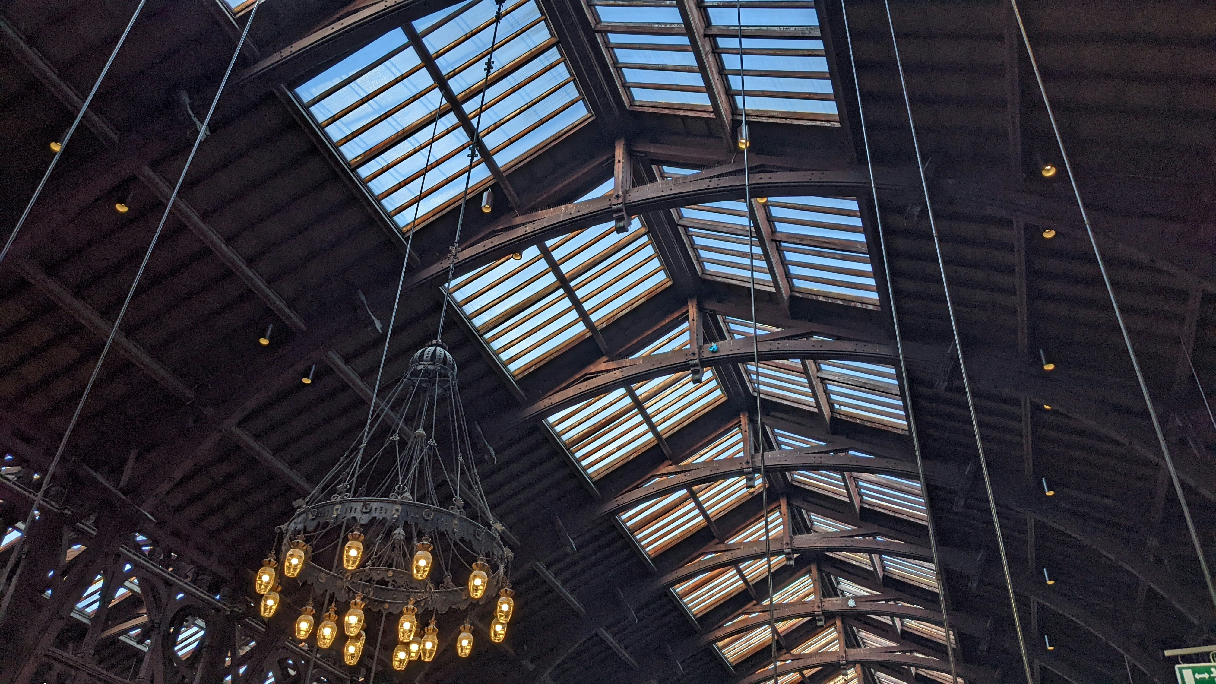 Exposed wooden beams and a vintage chandelier under a skylight in a spacious interior. The interplay of natural light and structural elements creates a warm atmosphere.