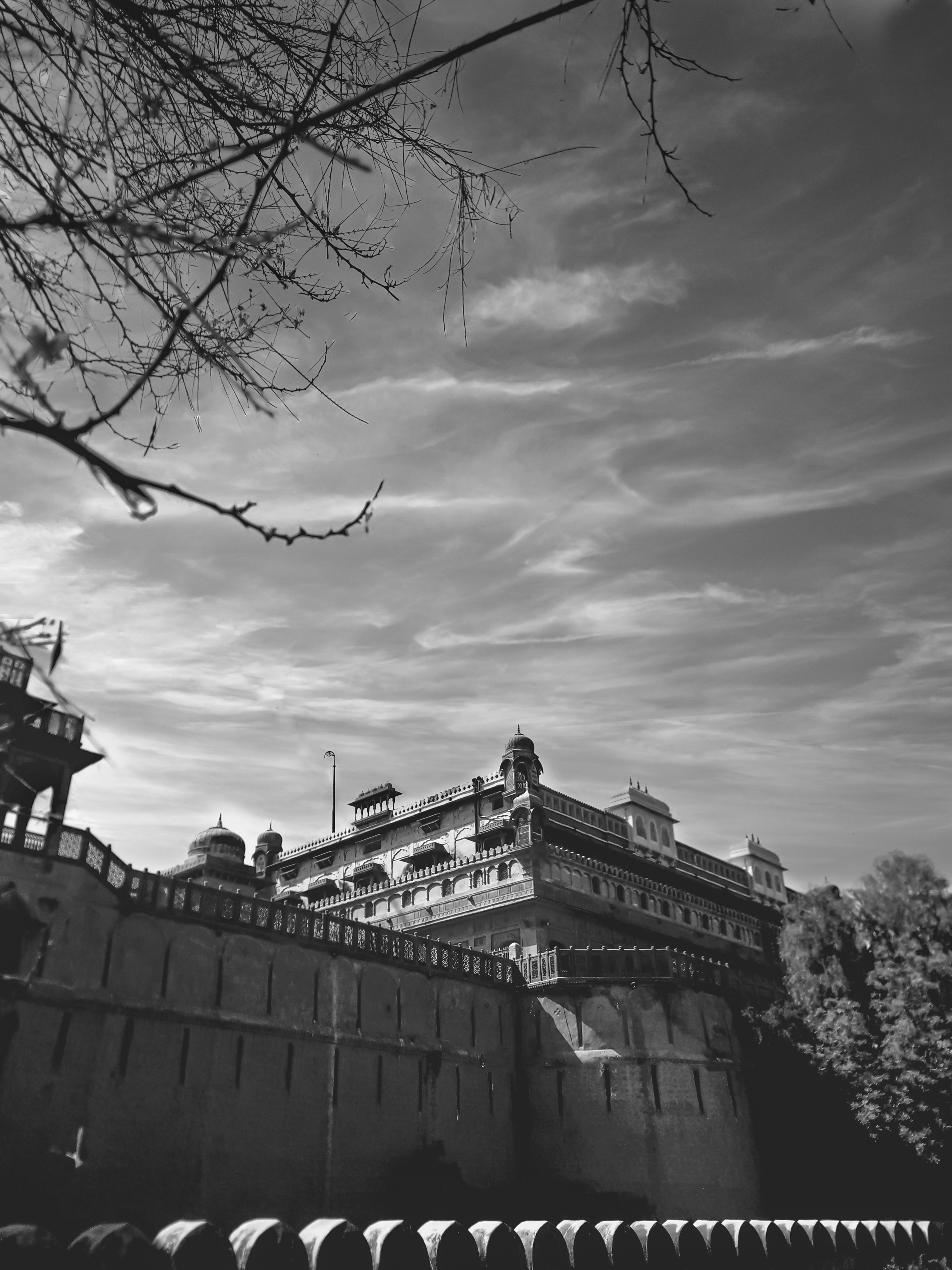 Black-and-white photograph of a fortress perched along a tall stone wall, framed by bare branches and a dramatic, cloud-filled sky.