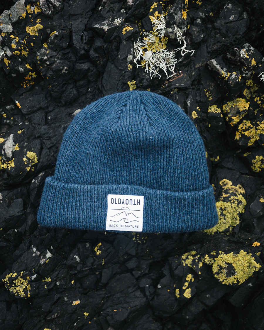 A blue knit beanie with a white logo patch rests on dark, textured volcanic rock dotted with yellow and white lichen. The clean product-style composition emphasizes the hat’s color and rugged outdoor setting.