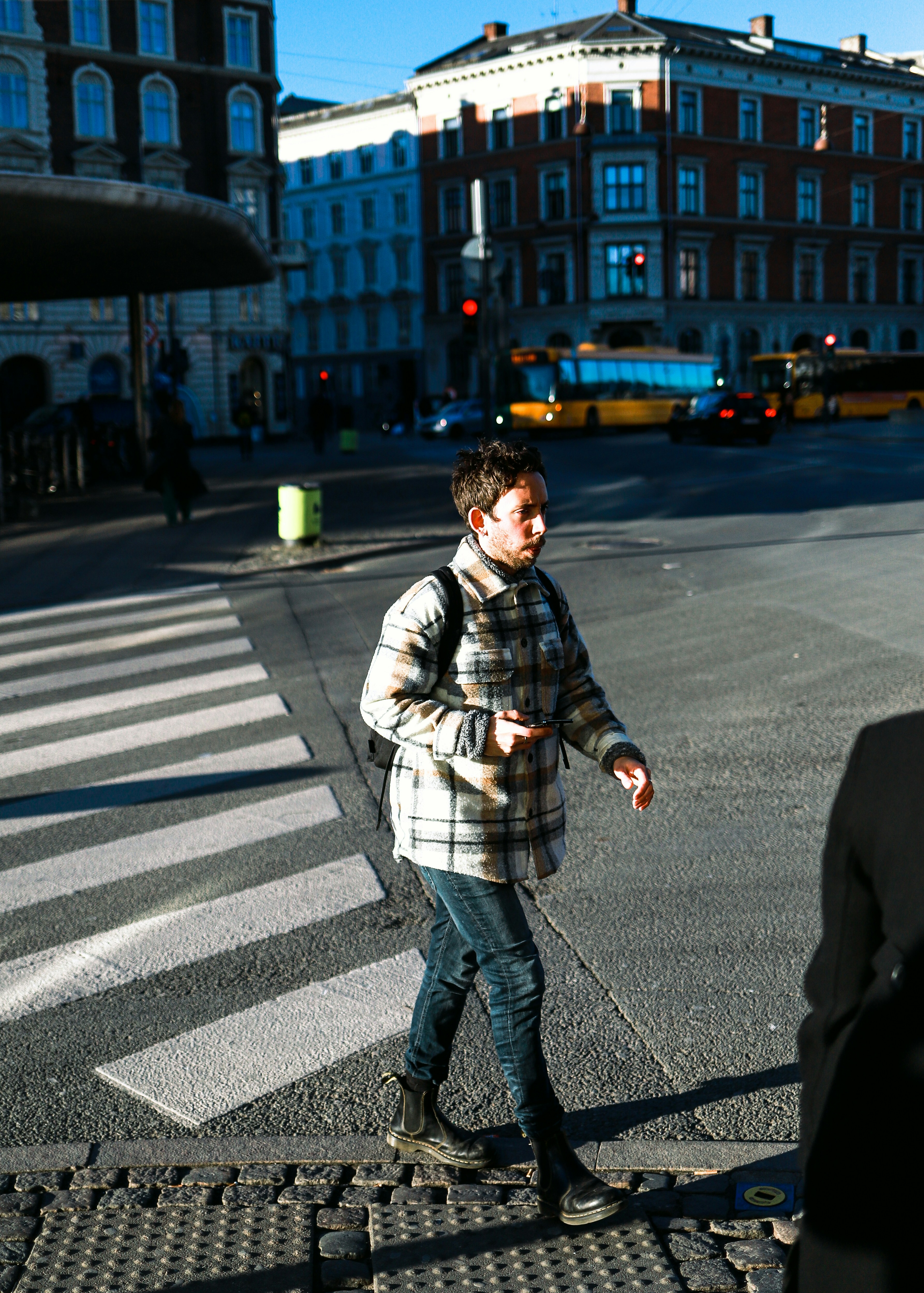 A pedestrian crosses the street, absorbed in their phone, amidst a bustling city backdrop featuring historical architecture and traffic lights.