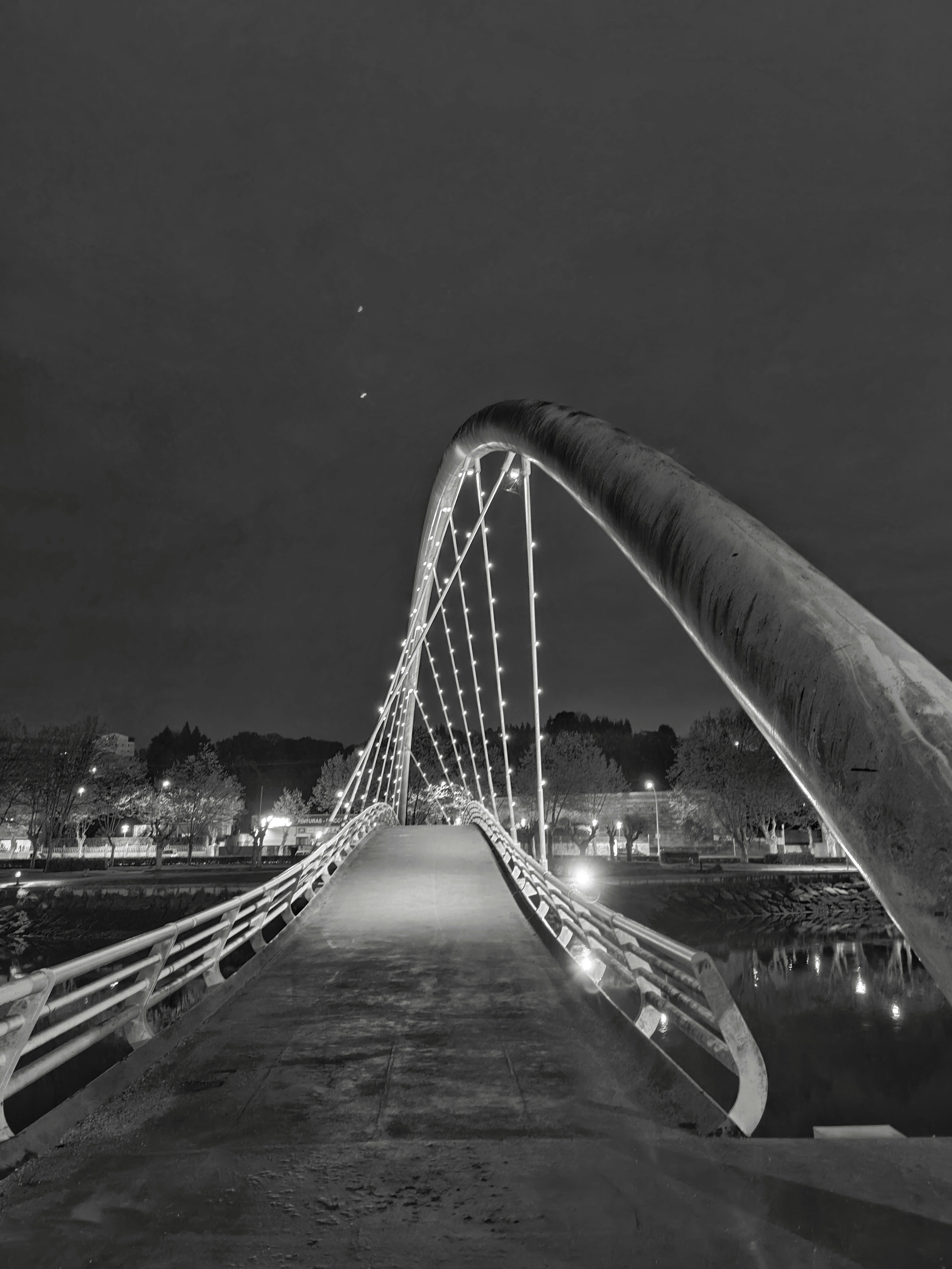 Illuminated pedestrian bridge gracefully arches over a calm waterway, surrounded by trees under a twilight sky.