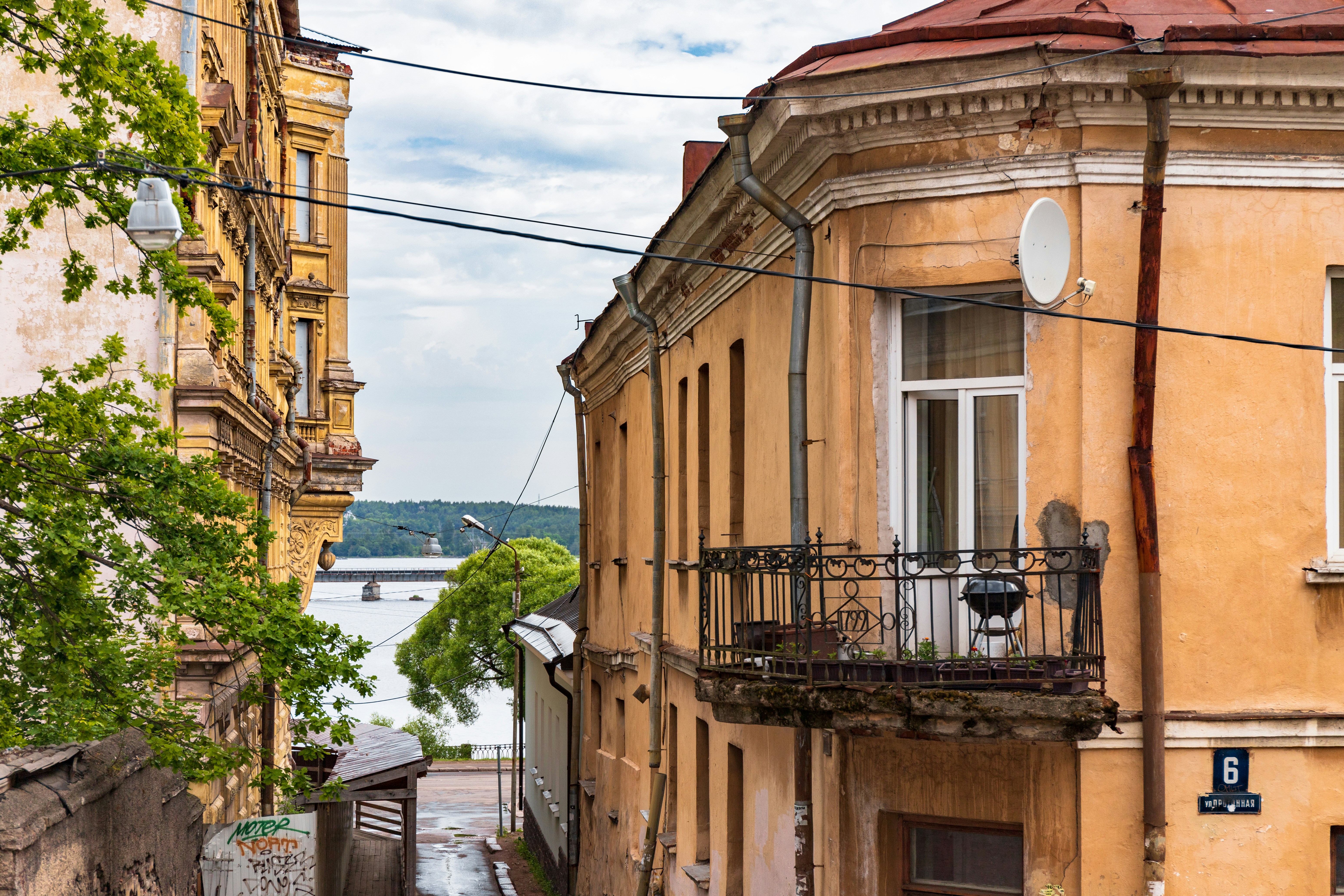 Narrow alley between aged buildings leading to a distant river under a cloudy sky.