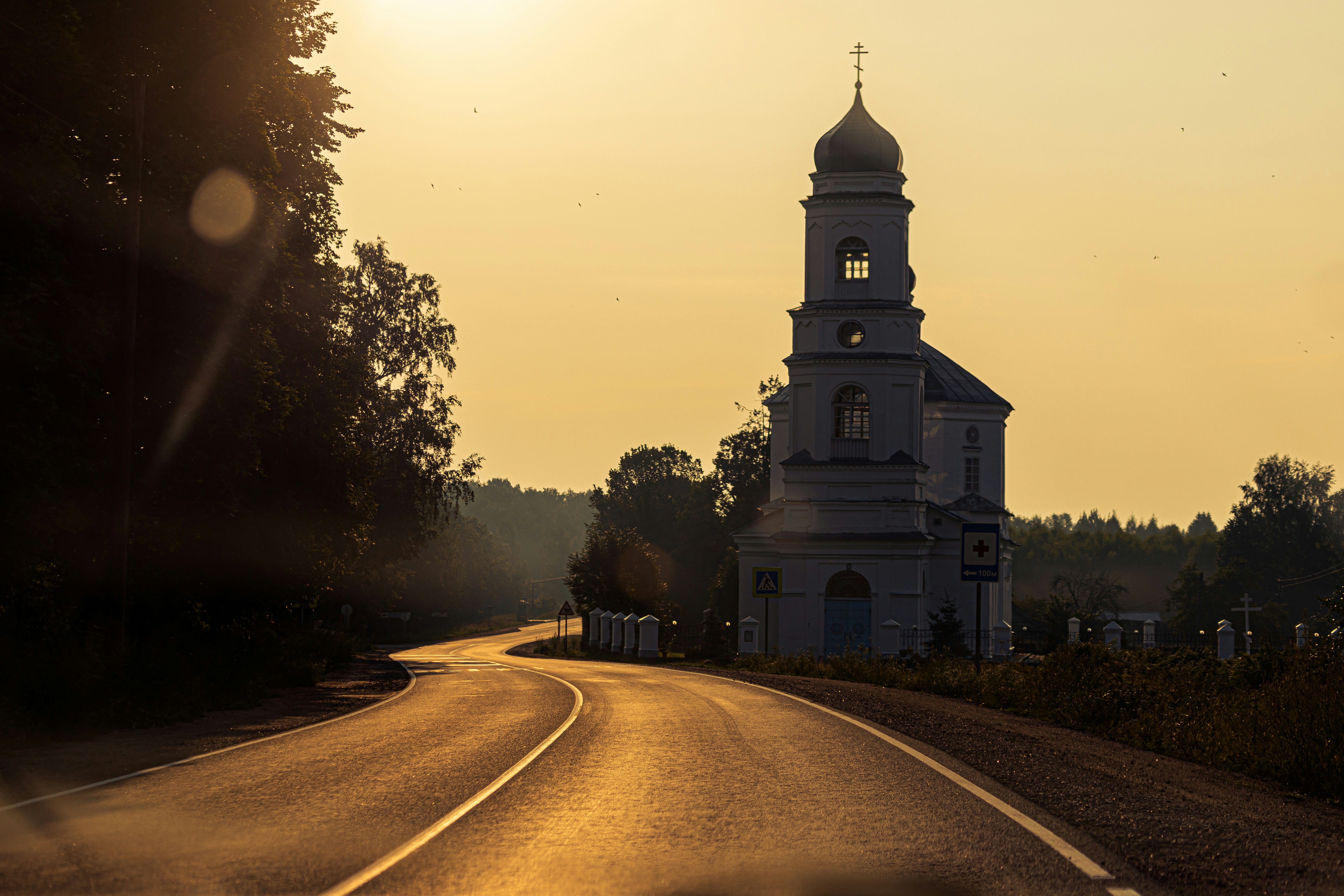 Sunlit rural road curving towards a solitary chapel at sunset.