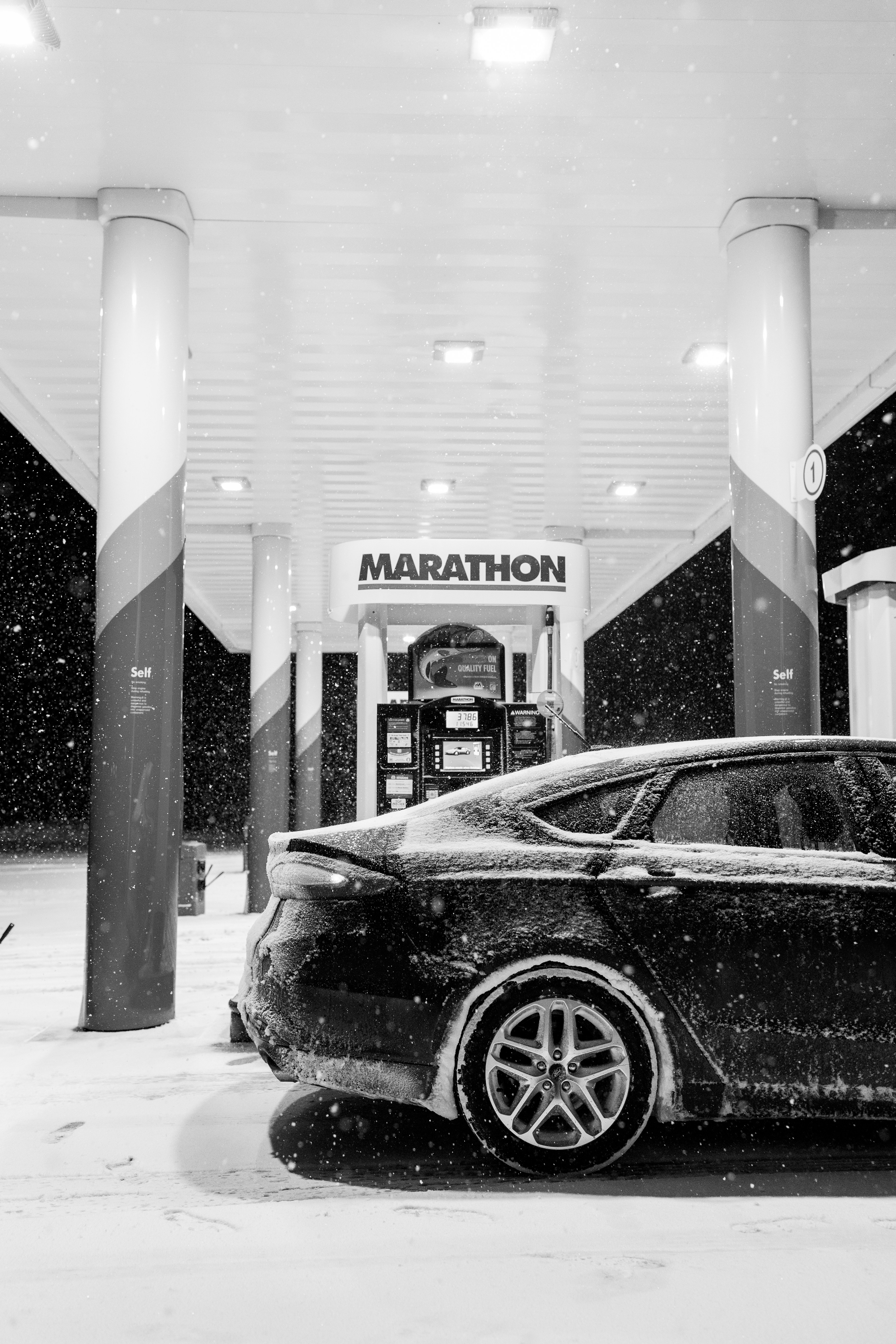 Snow-covered car parked at a Marathon gas station during a snowfall at night.
