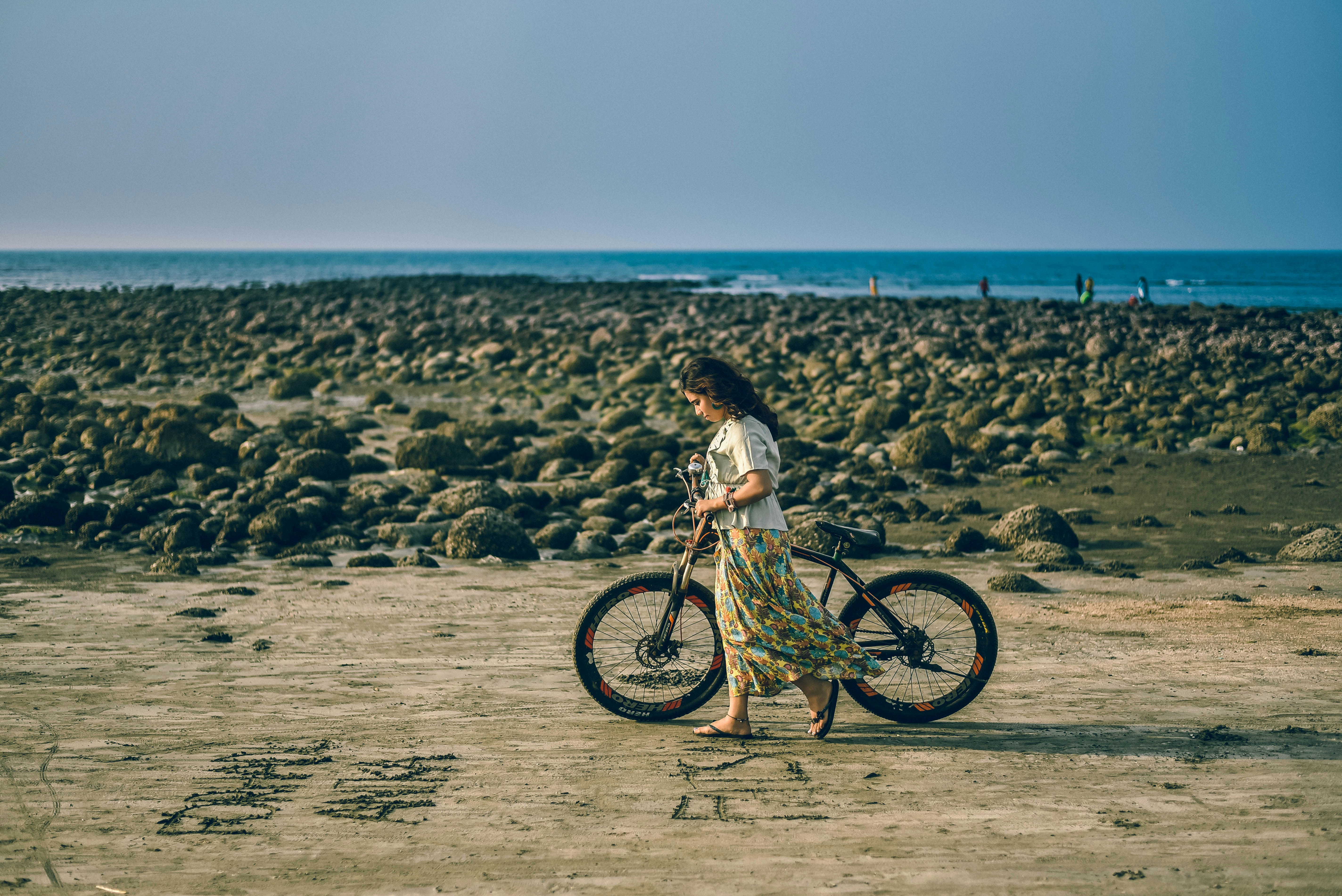 M road-walking along the shore: woman with bicycle by rocky beach