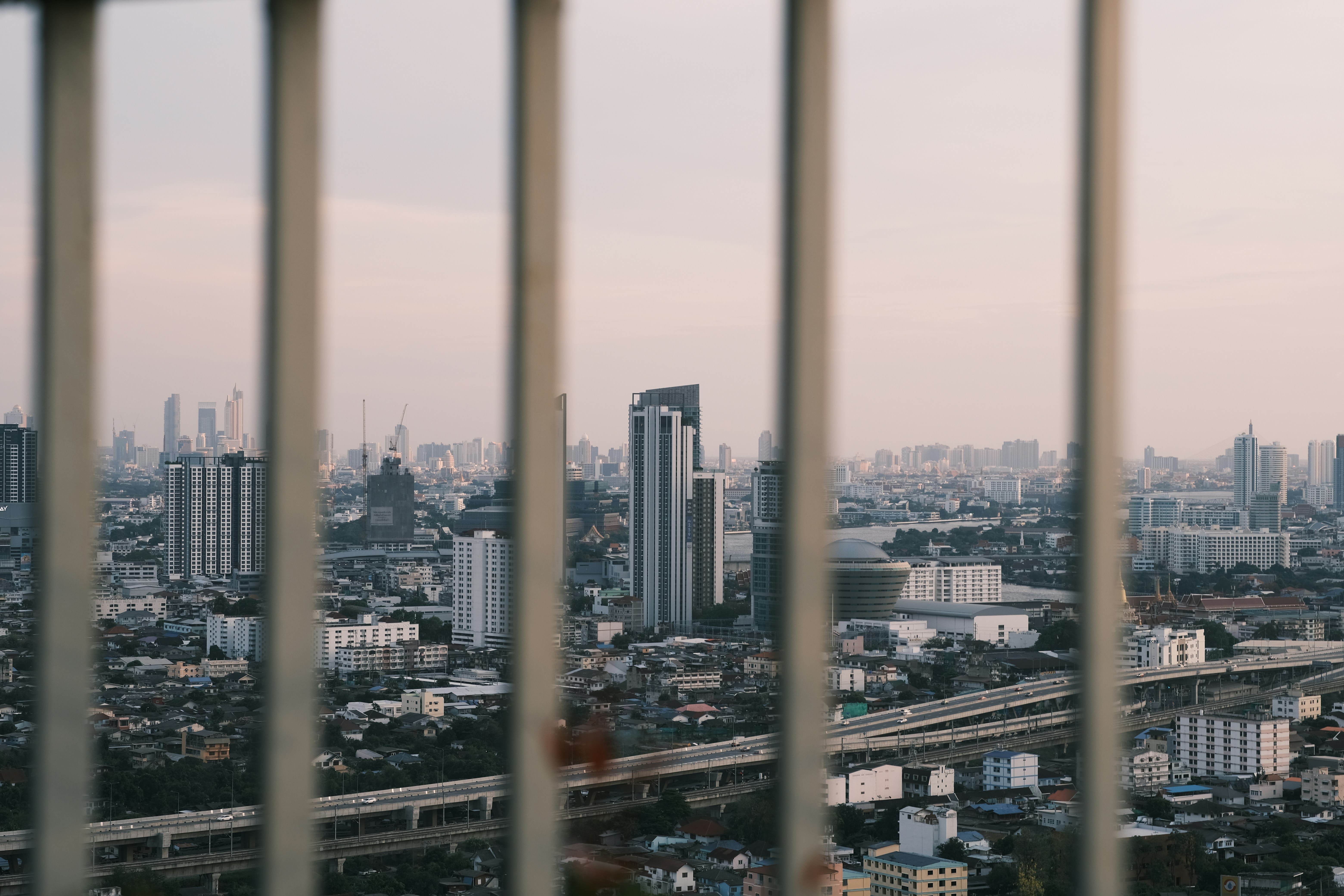 A panoramic view of a bustling city skyline captured through the bars of a railing, highlighting urban architecture and a hint of nature below.