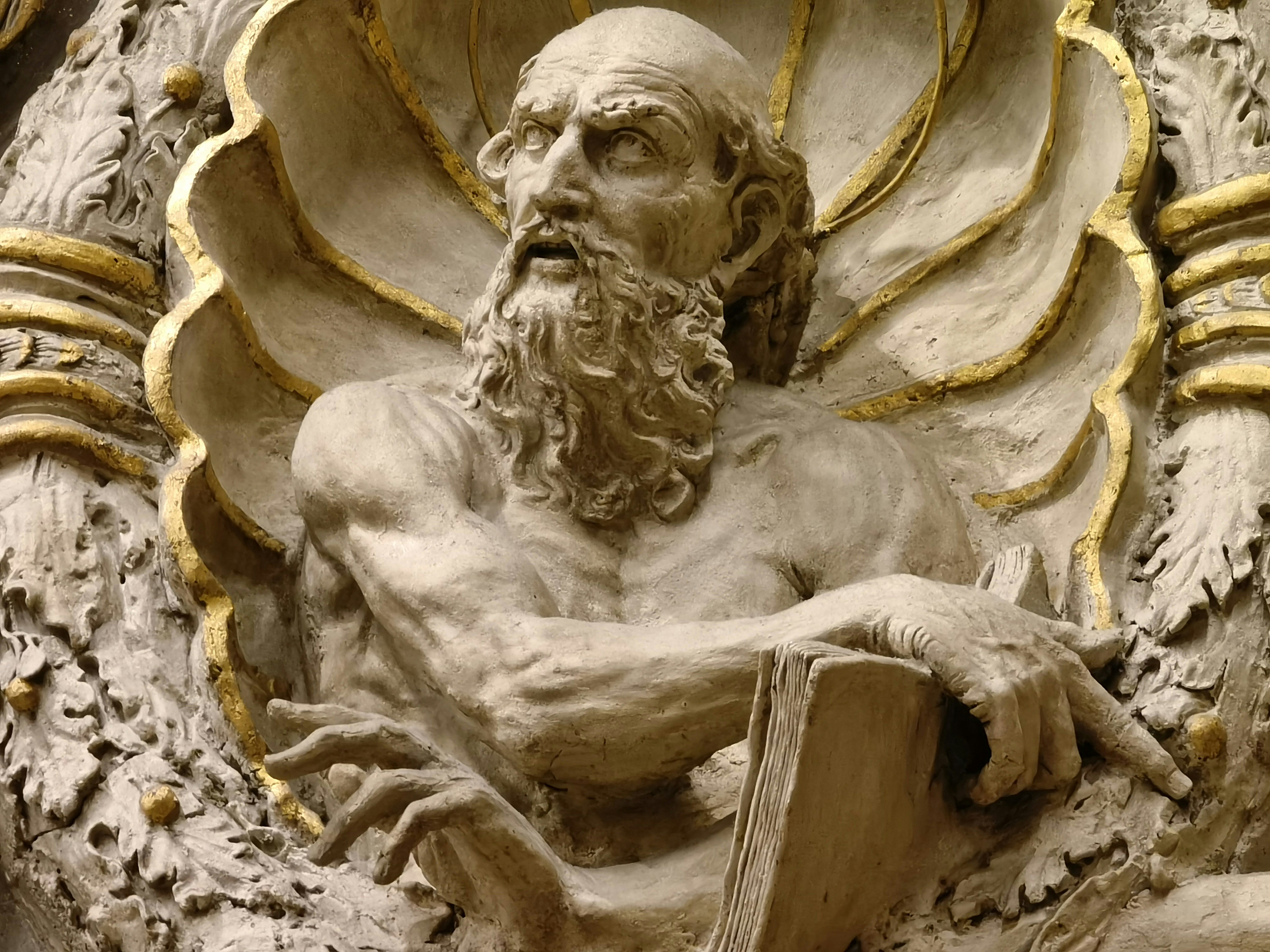 Intricately detailed stone sculpture of a bearded figure holding a book, set against a shell-like backdrop with golden accents.