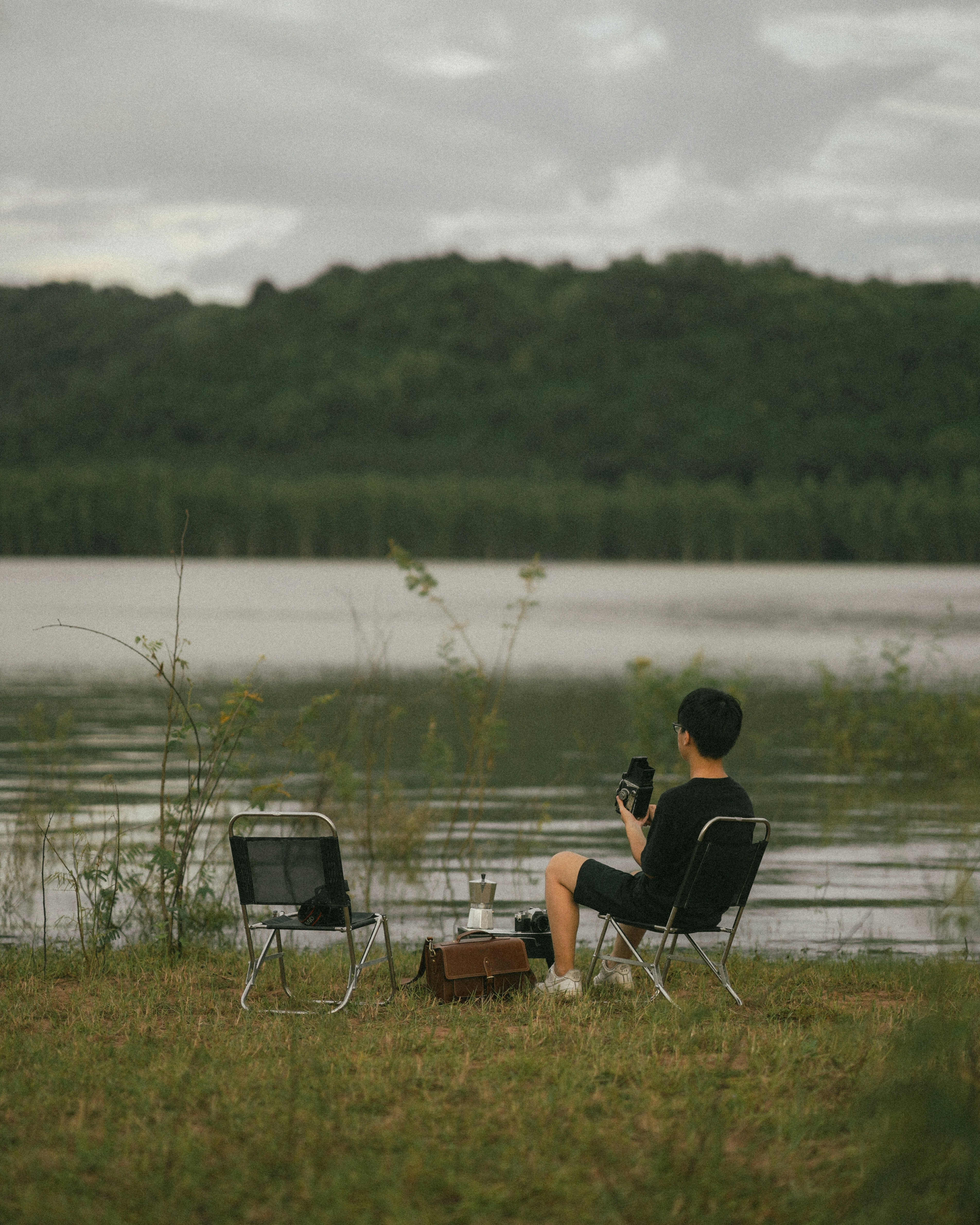 A person seated on a chair by the lake, holding a camera, surrounded by nature. The calm water reflects the lush greenery in the background.
