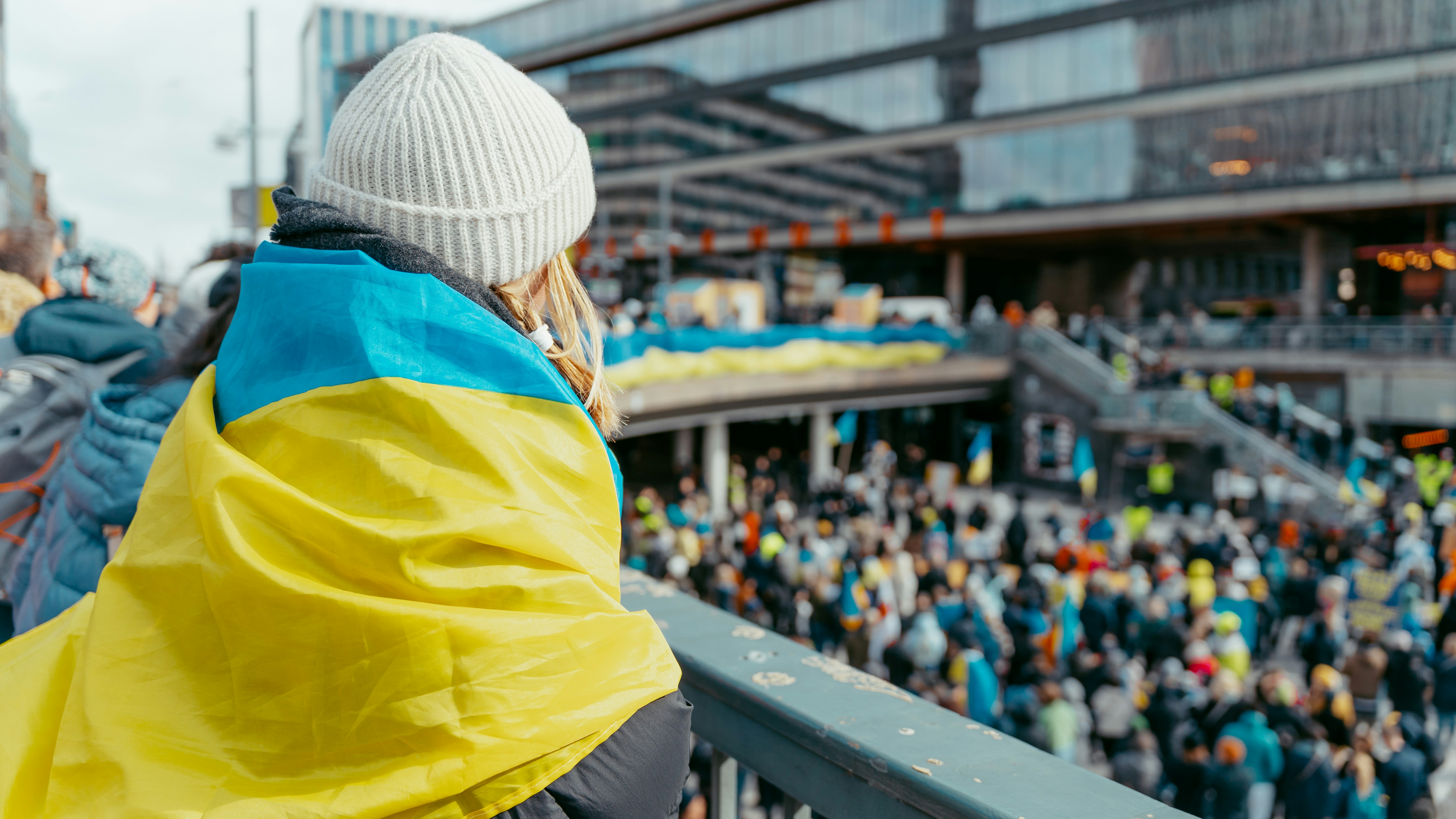 Individual wrapped in a Ukrainian flag observes a large gathering, symbolizing solidarity and support. The urban backdrop enhances the communal atmosphere.