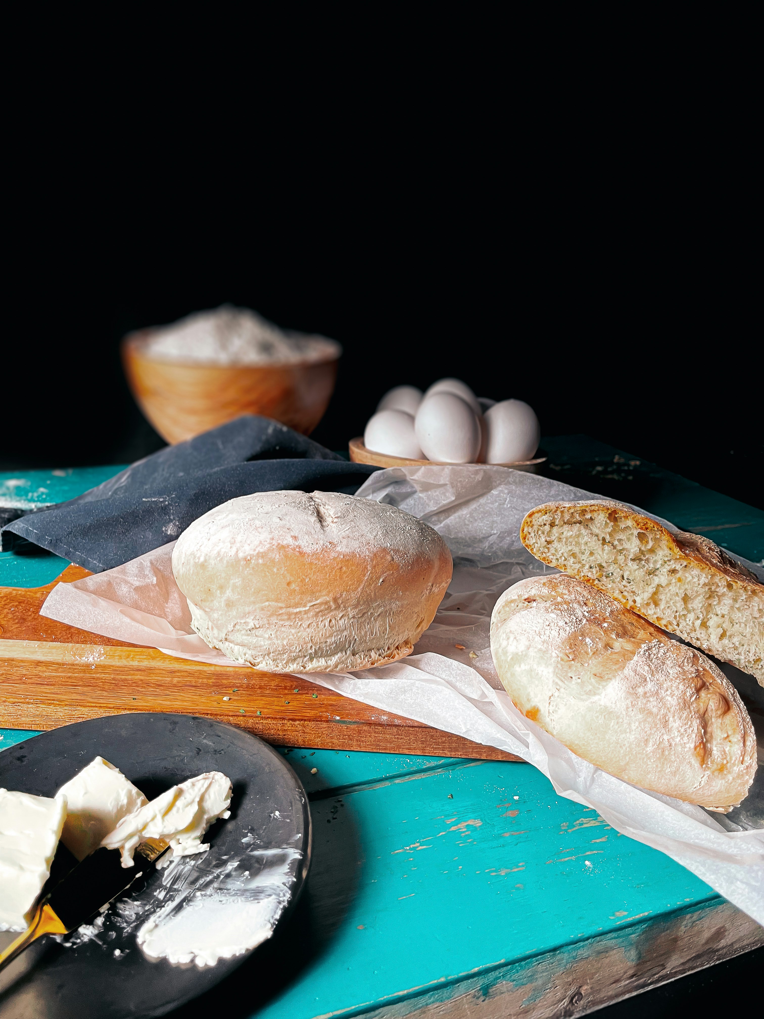 Freshly baked artisan bread rests on a wooden cutting board alongside butter and eggs, with a bowl of flour in the background. The dark backdrop emphasizes the textures and colors of the ingredients.