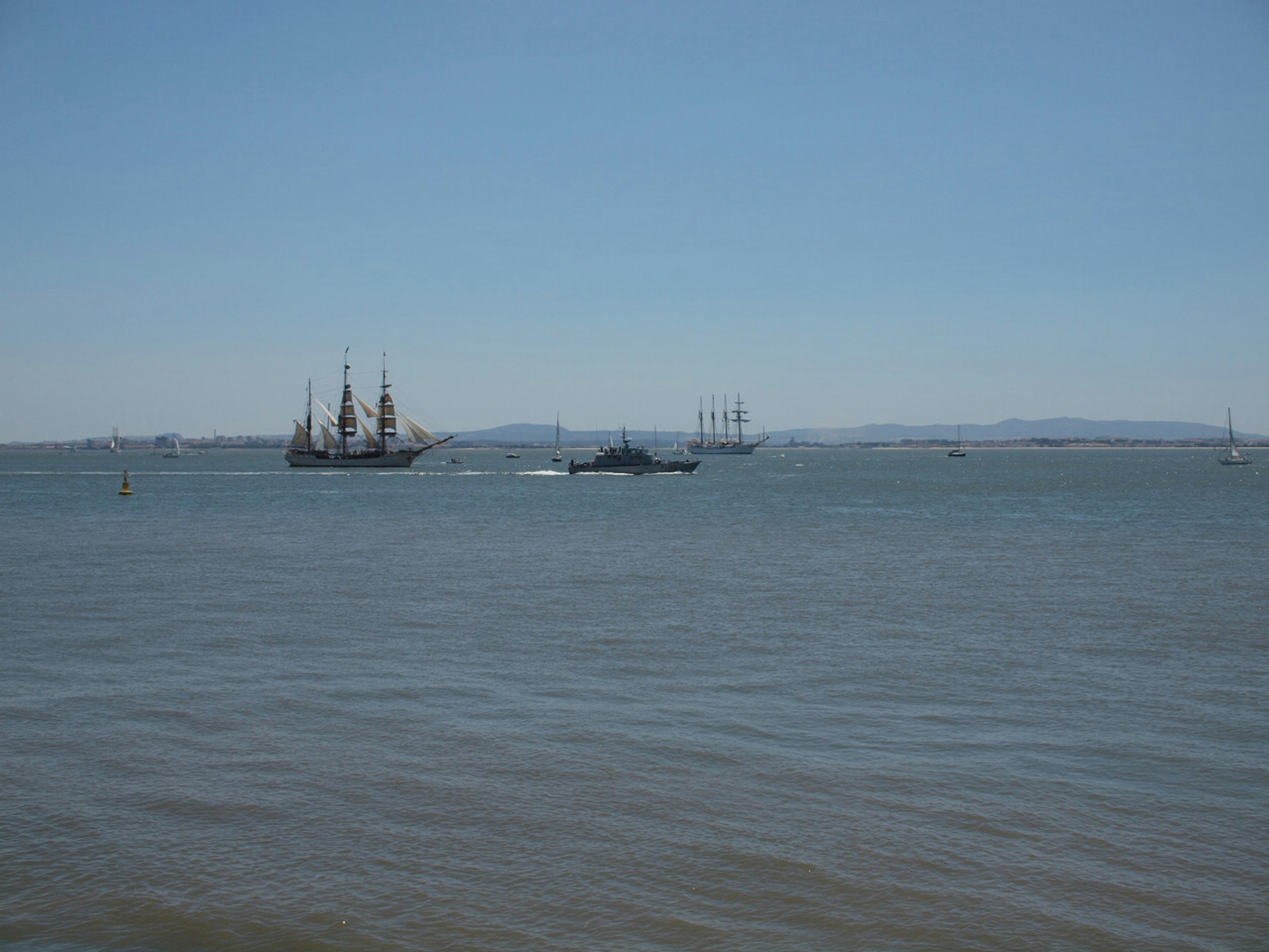 Calm coastal water with several sailing ships near the horizon and a distant shoreline; appears to be a harbor scene with a small buoy in the foreground. A simple, tranquil maritime view.