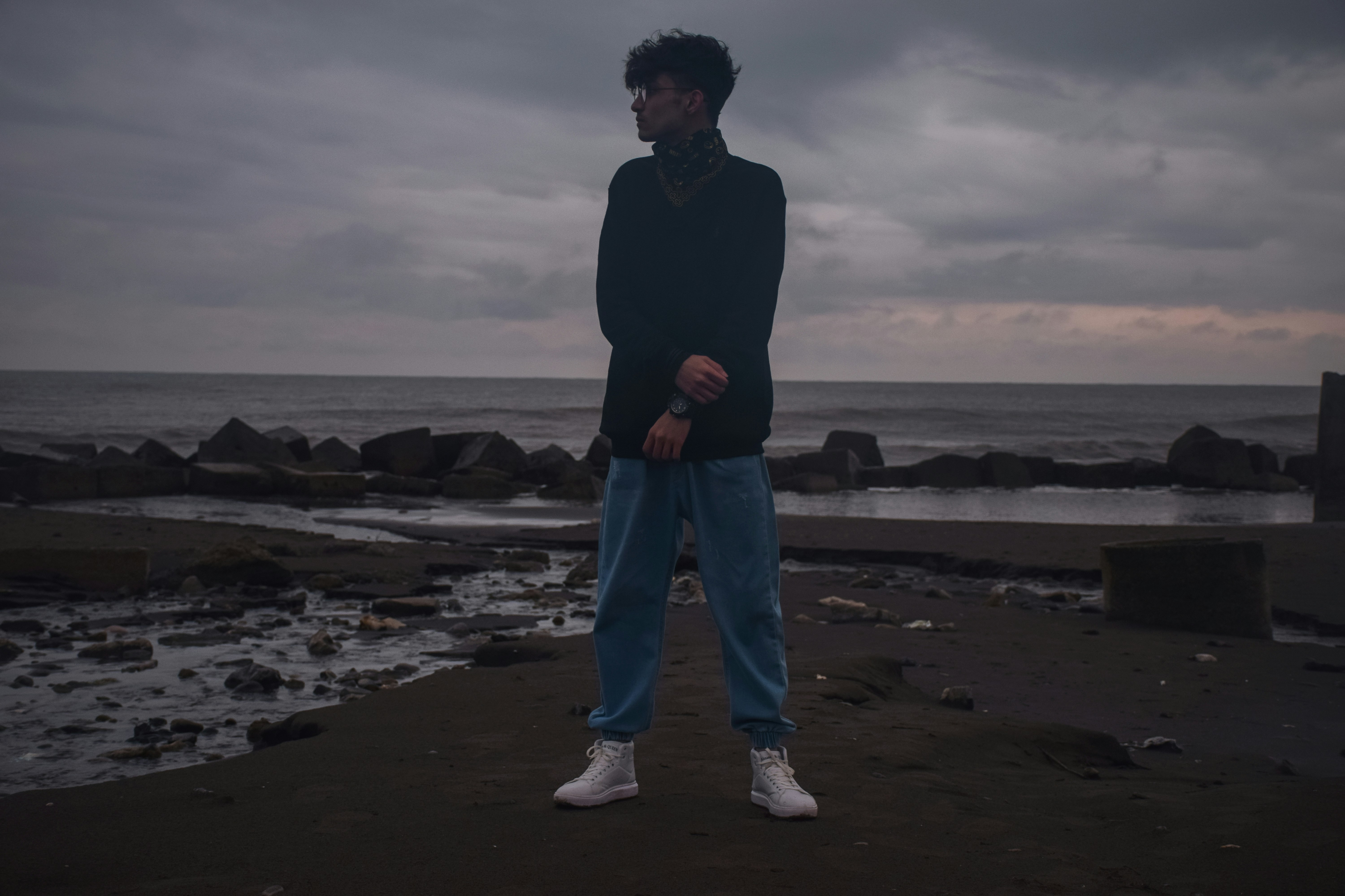 Person standing on a dimly lit beach with a cloudy sky and calm sea in the background.