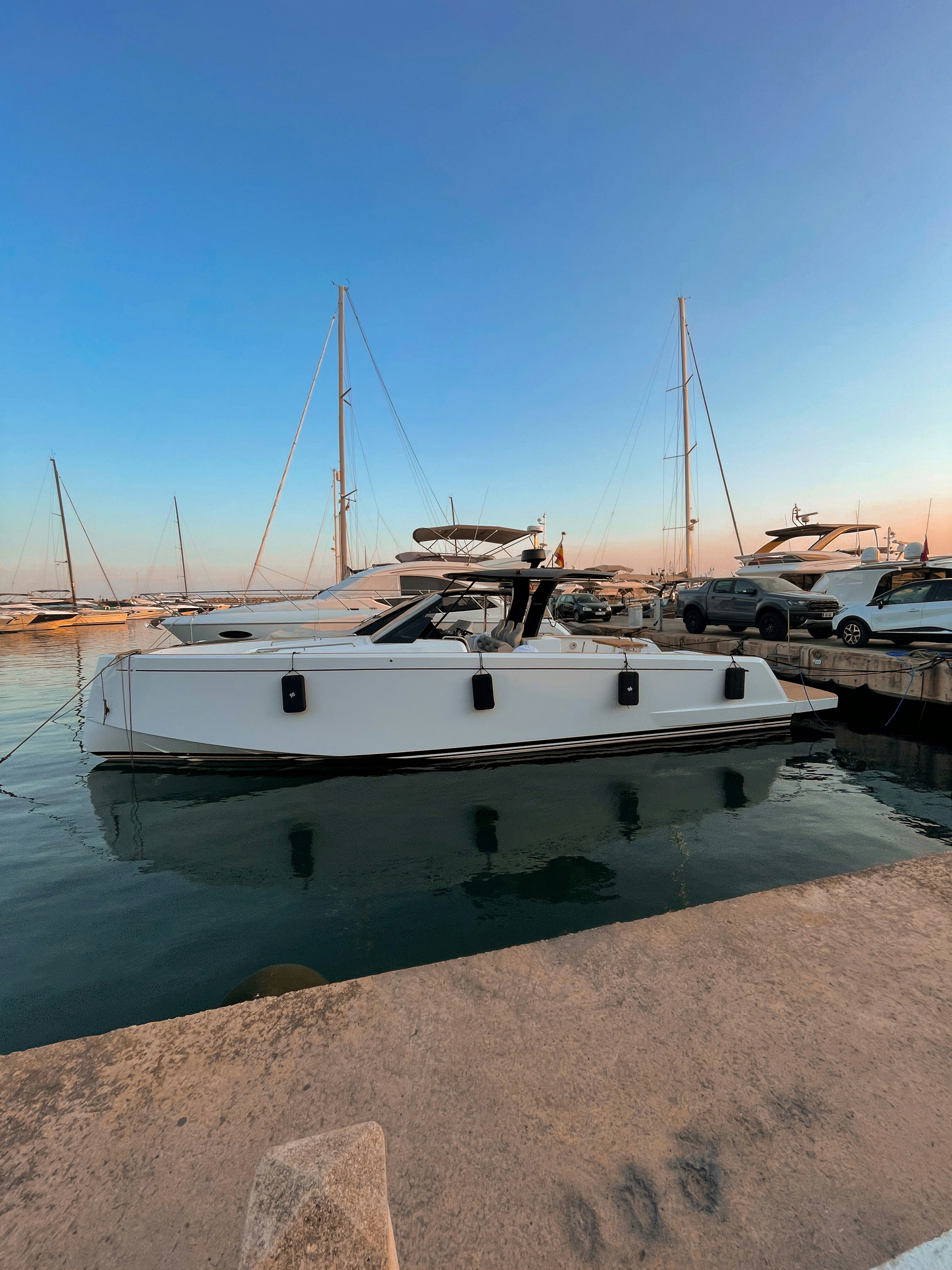 White yacht docked at a marina during sunset with sailboats in the background and a clear sky overhead.