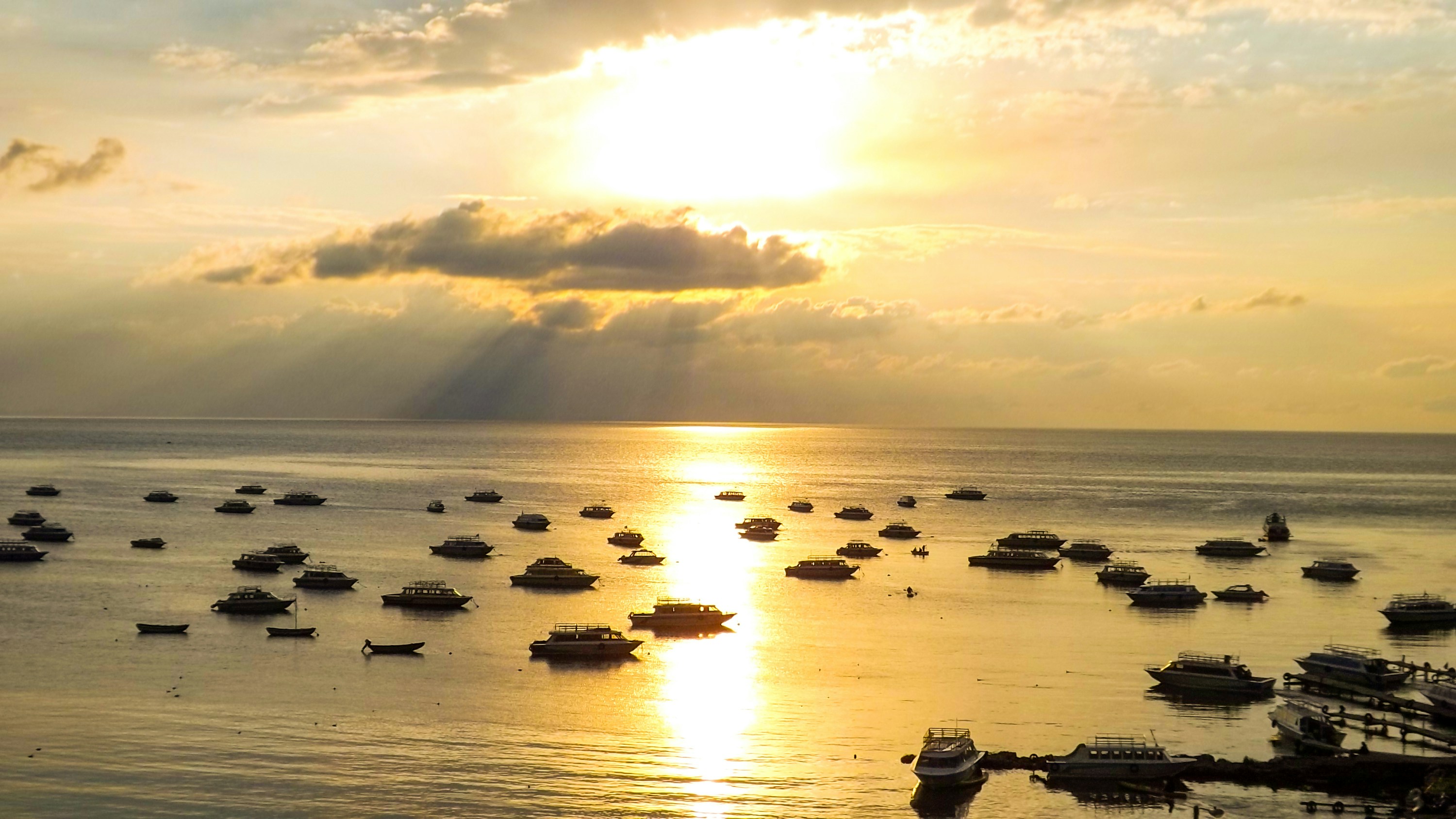 Boats scattered across a calm sea under a golden sunrise, with sunbeams piercing through clouds.