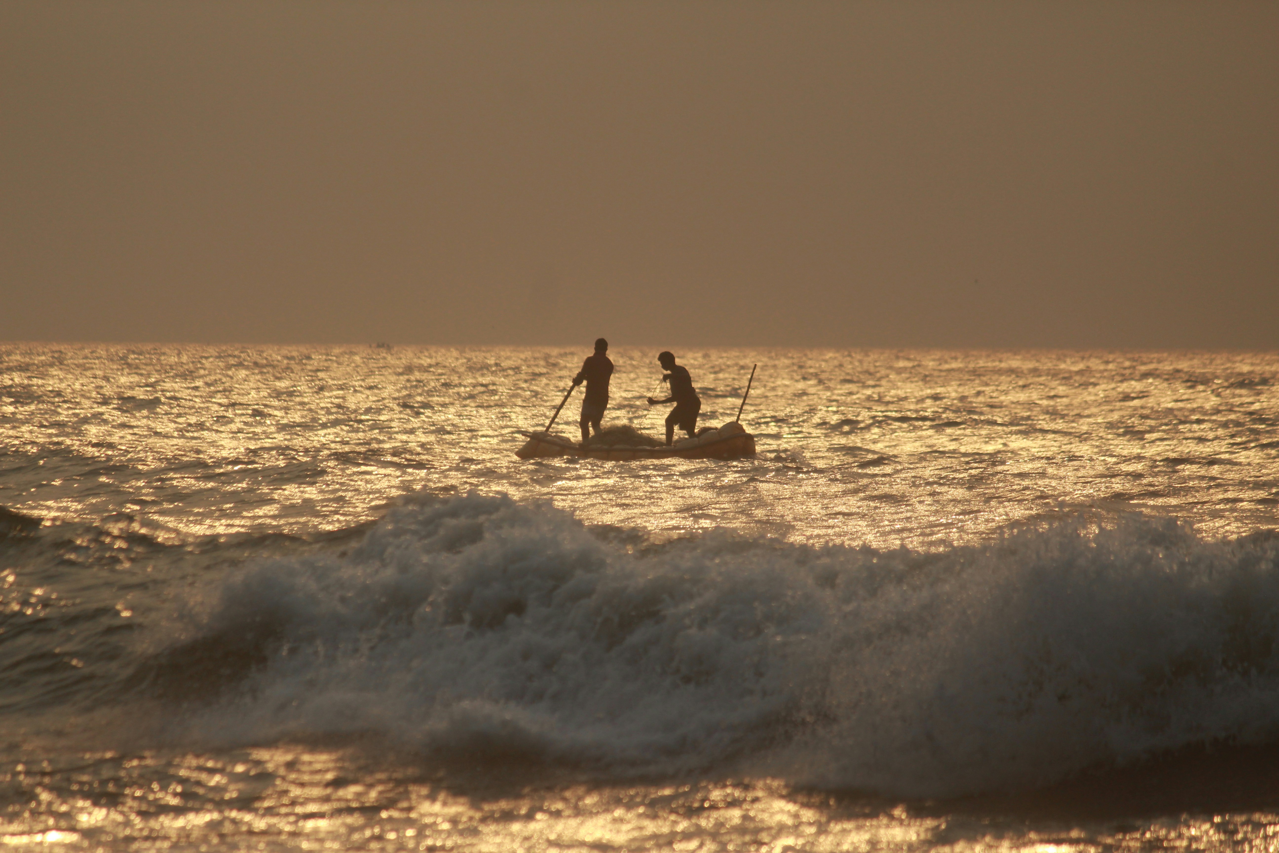Two fishermen navigating their boat through glistening waves at sunrise, surrounded by a shimmering ocean surface.