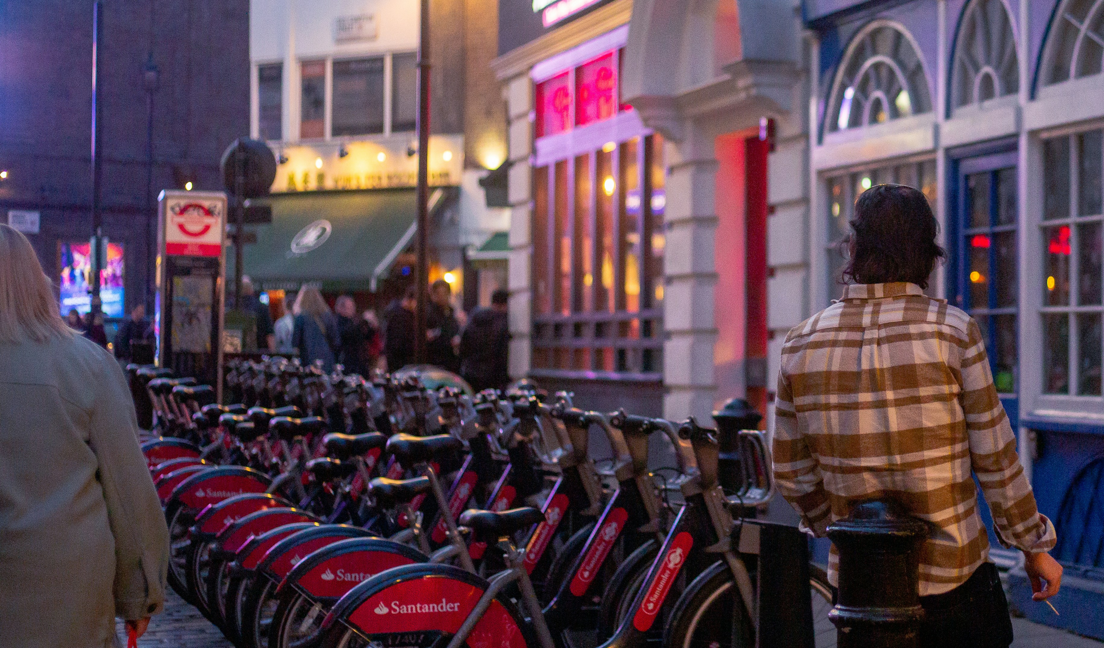 A bustling urban street scene featuring a row of Santander bicycles against a backdrop of vibrant nightlife, with pedestrians enjoying the evening ambiance.