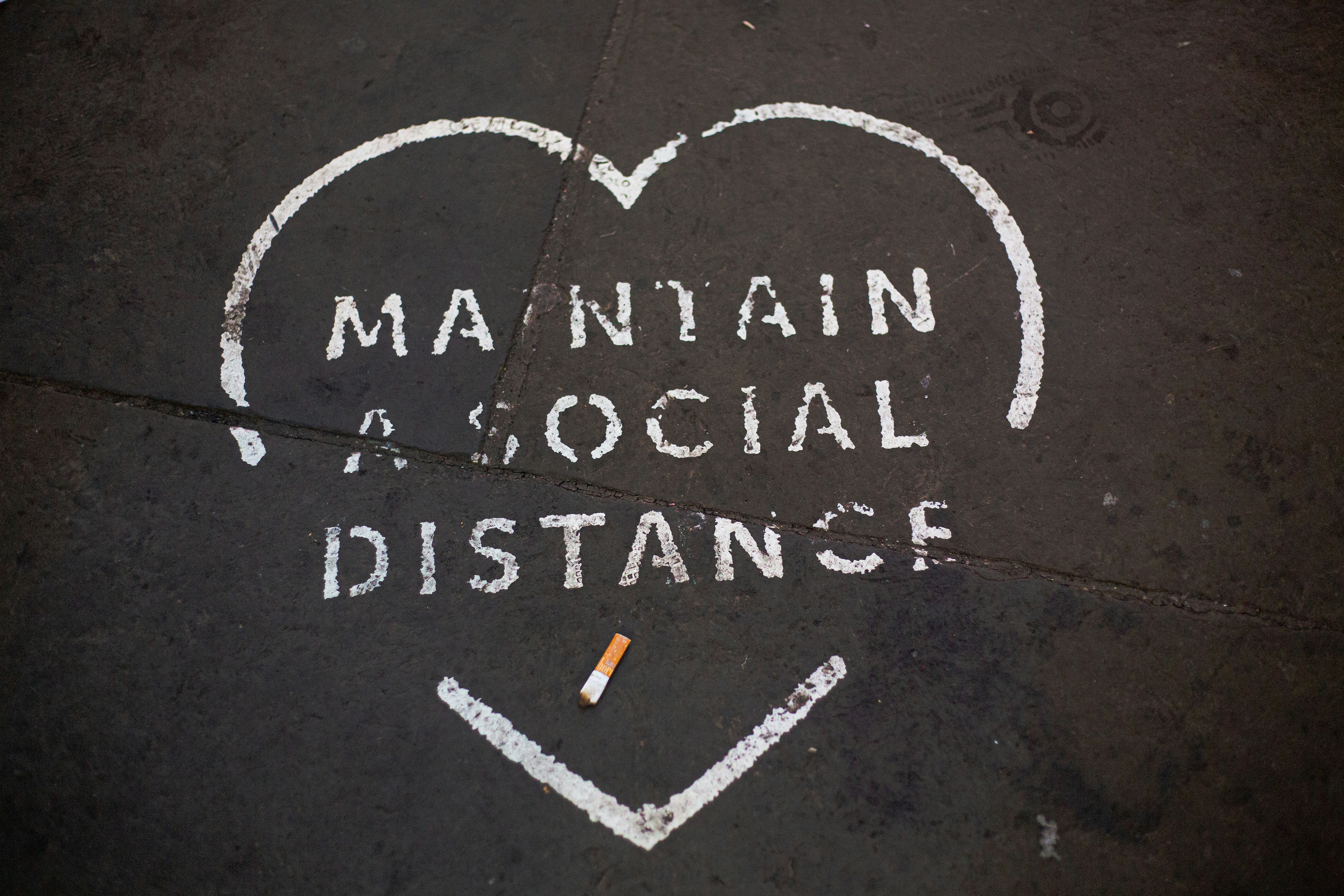 White chalk lettering urging to maintain social distance inside a heart shape on the ground, accompanied by a discarded cigarette. 