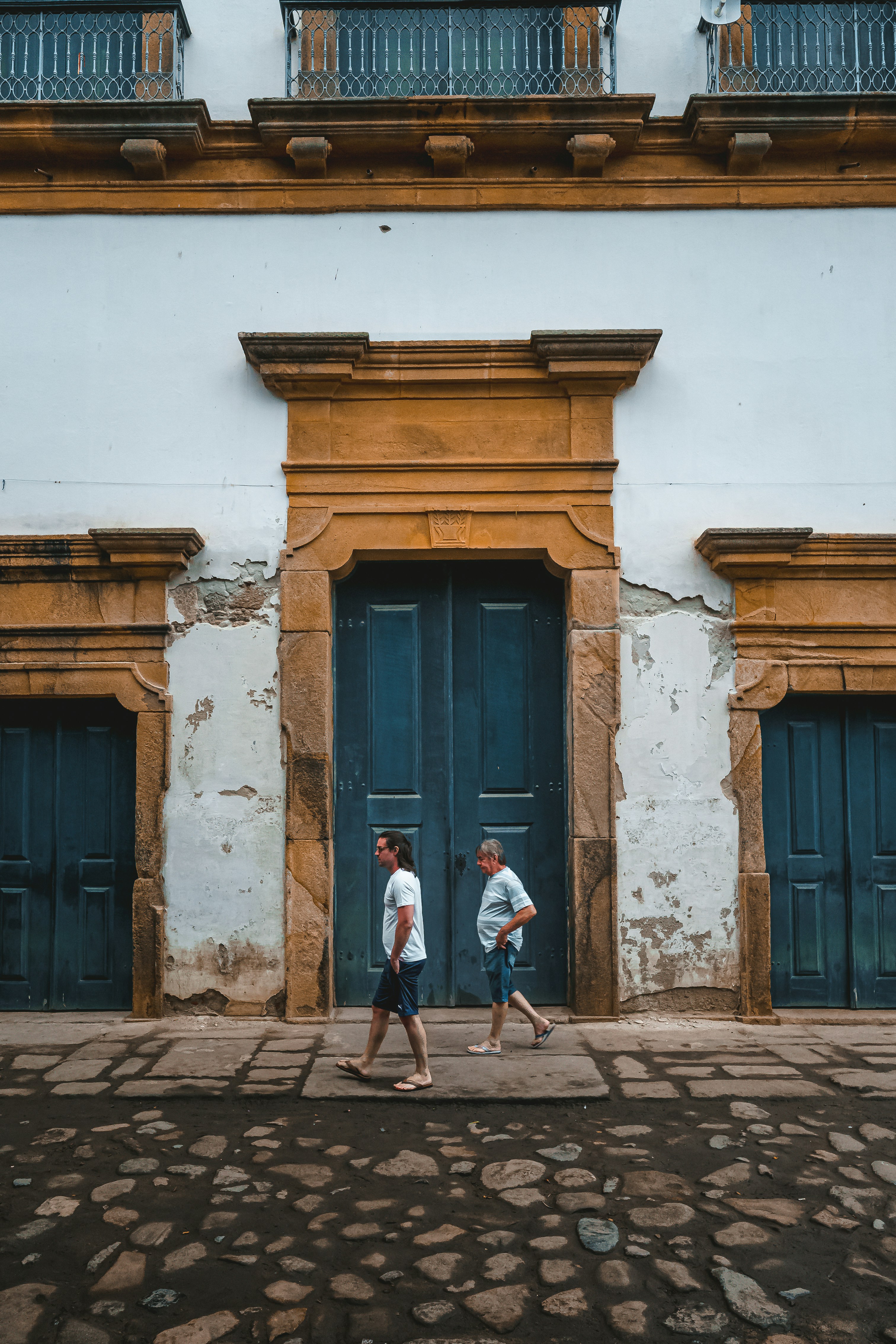 Two individuals walking past a weathered building with blue doors and textured walls, showcasing a blend of history and everyday life.