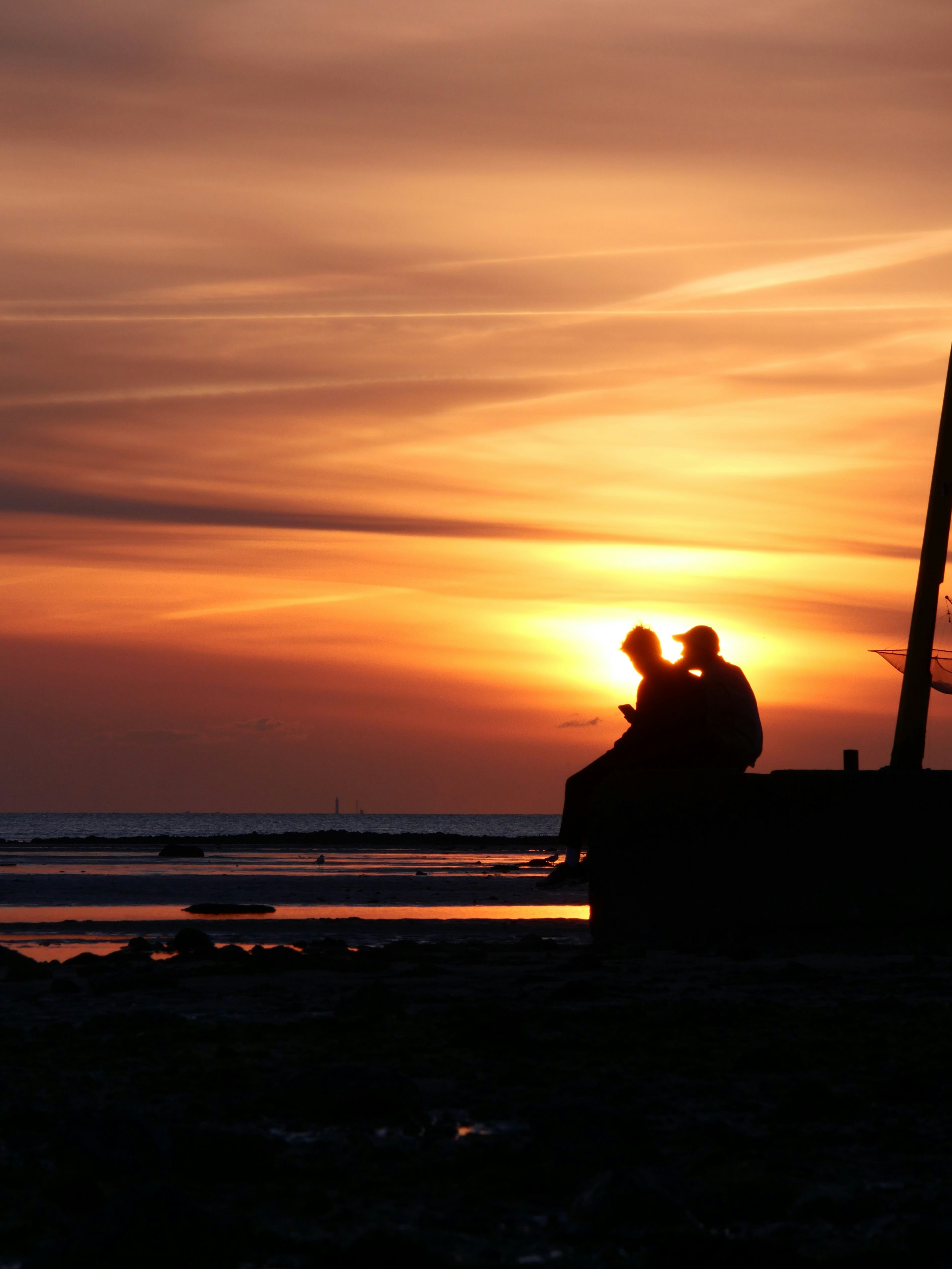 Two silhouetted figures sit close on a railing by the shoreline as a warm sunset floods the horizon.