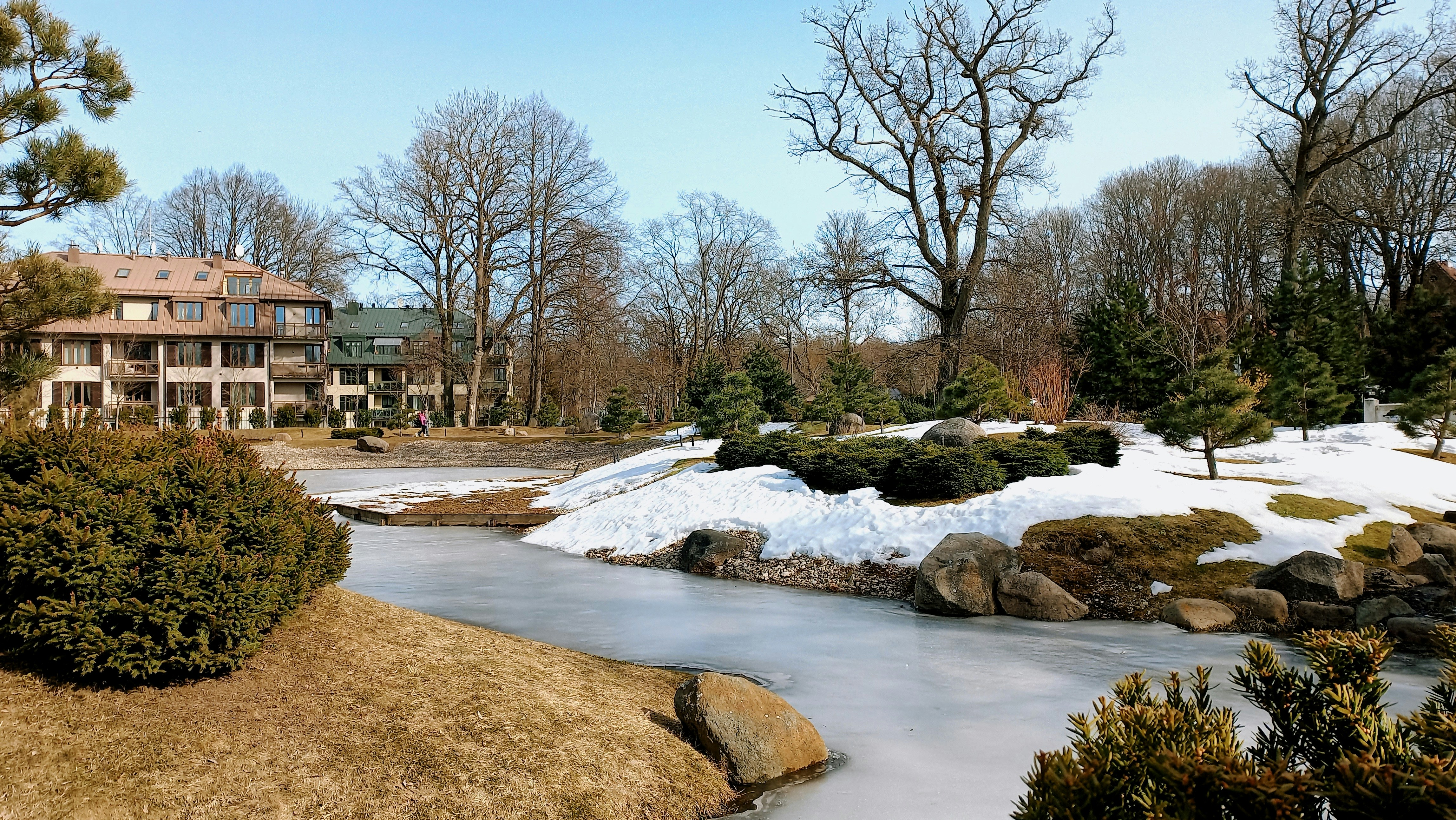 Tranquil landscape featuring a frozen stream bordered by snow and greenery, with a modern building in the background. The scene captures the peaceful coexistence of winter and early spring.