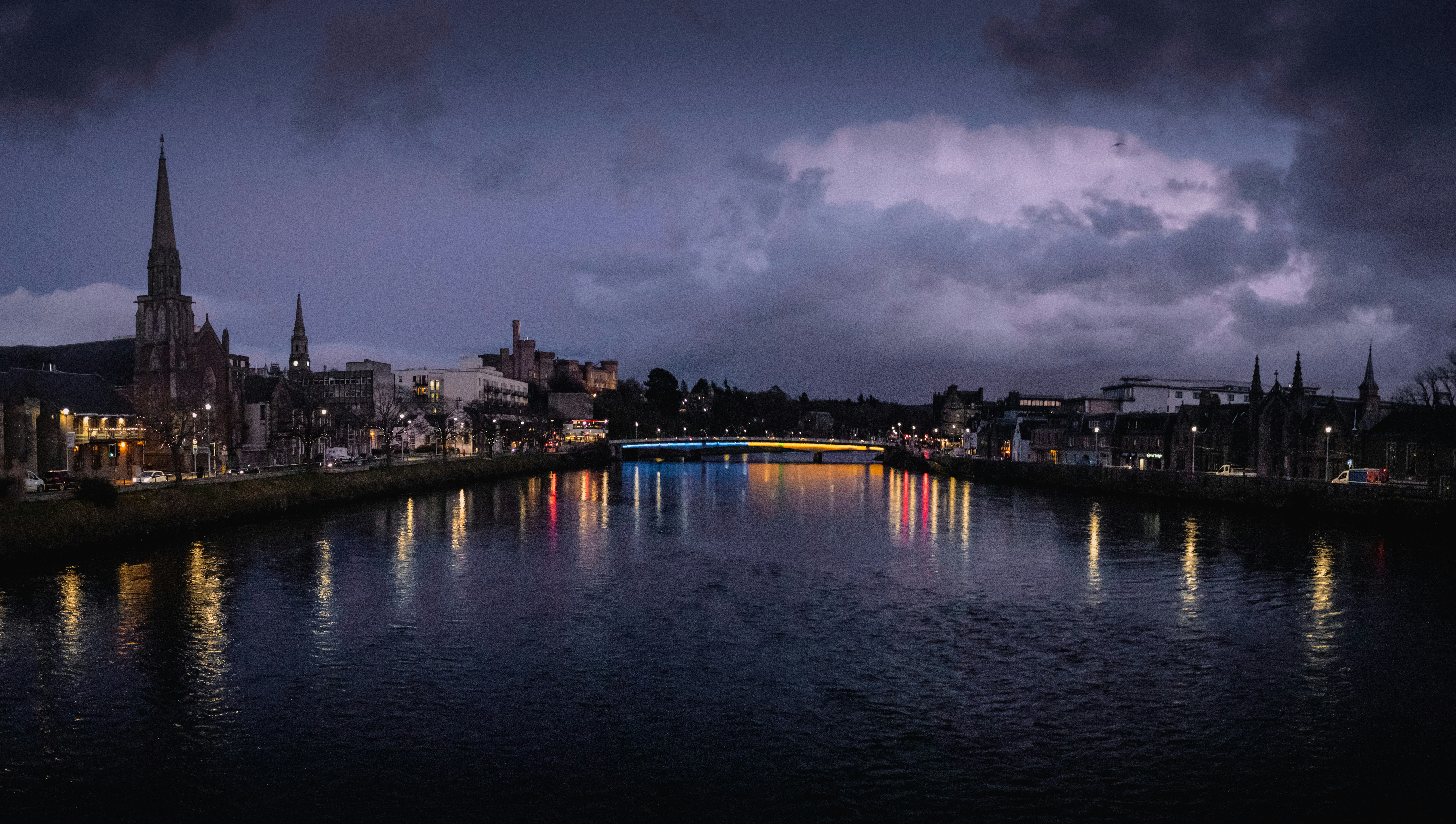Evening view of Inverness cityscape with illuminated buildings reflecting on the River Ness under a moody sky.