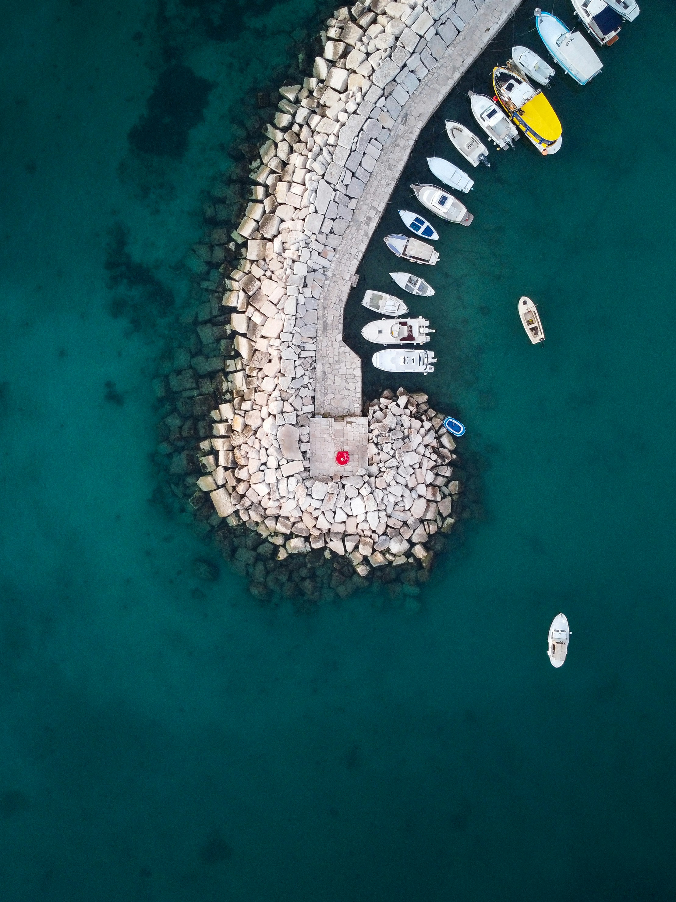 Aerial view of a stone pier with boats moored alongside, featuring a vibrant red umbrella at the end. The clear turquoise water enhances the tranquil atmosphere.