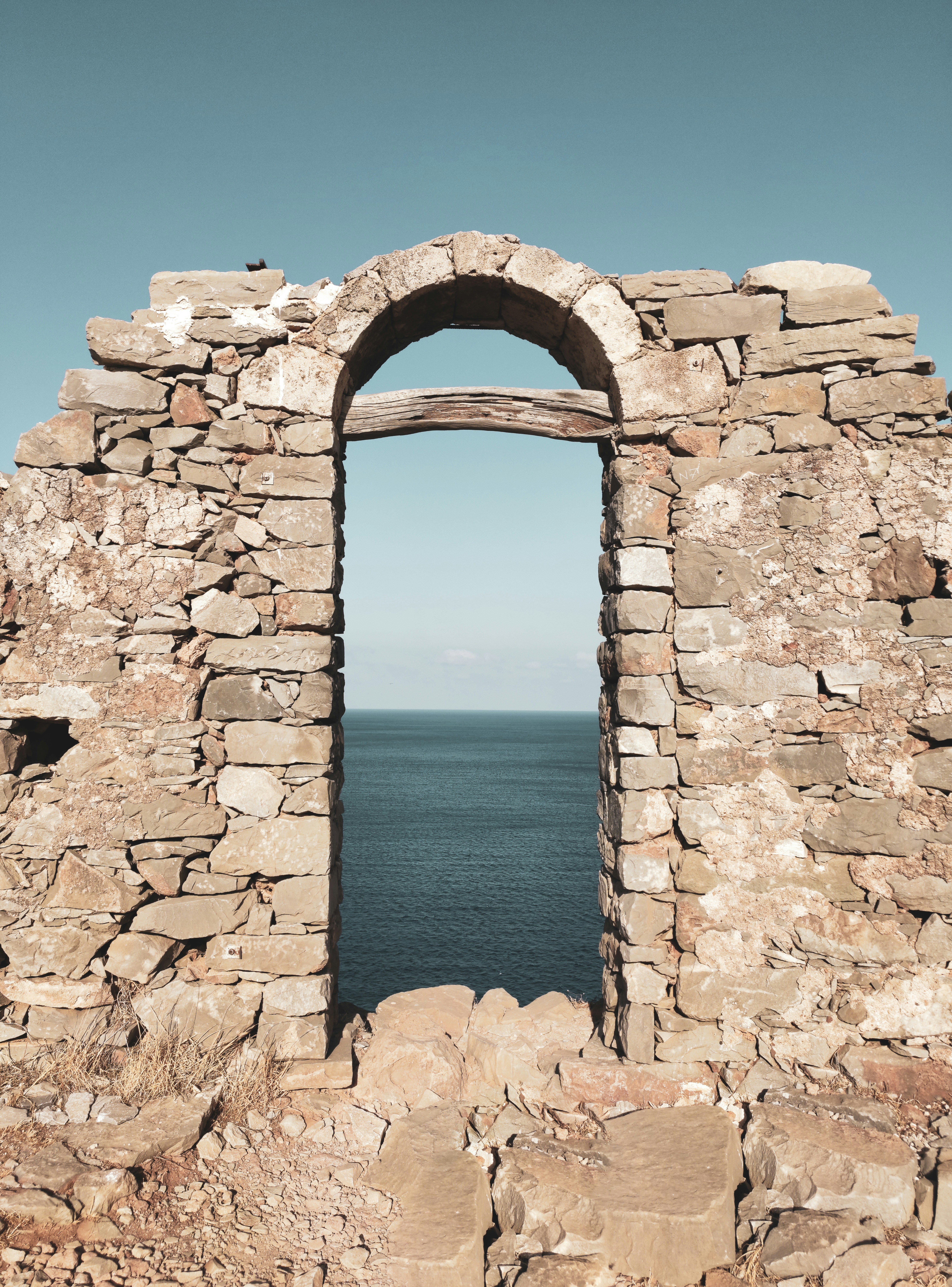Weathered stone archway overlooking a tranquil sea under a clear sky. The remnants of the structure hint at a rich history.