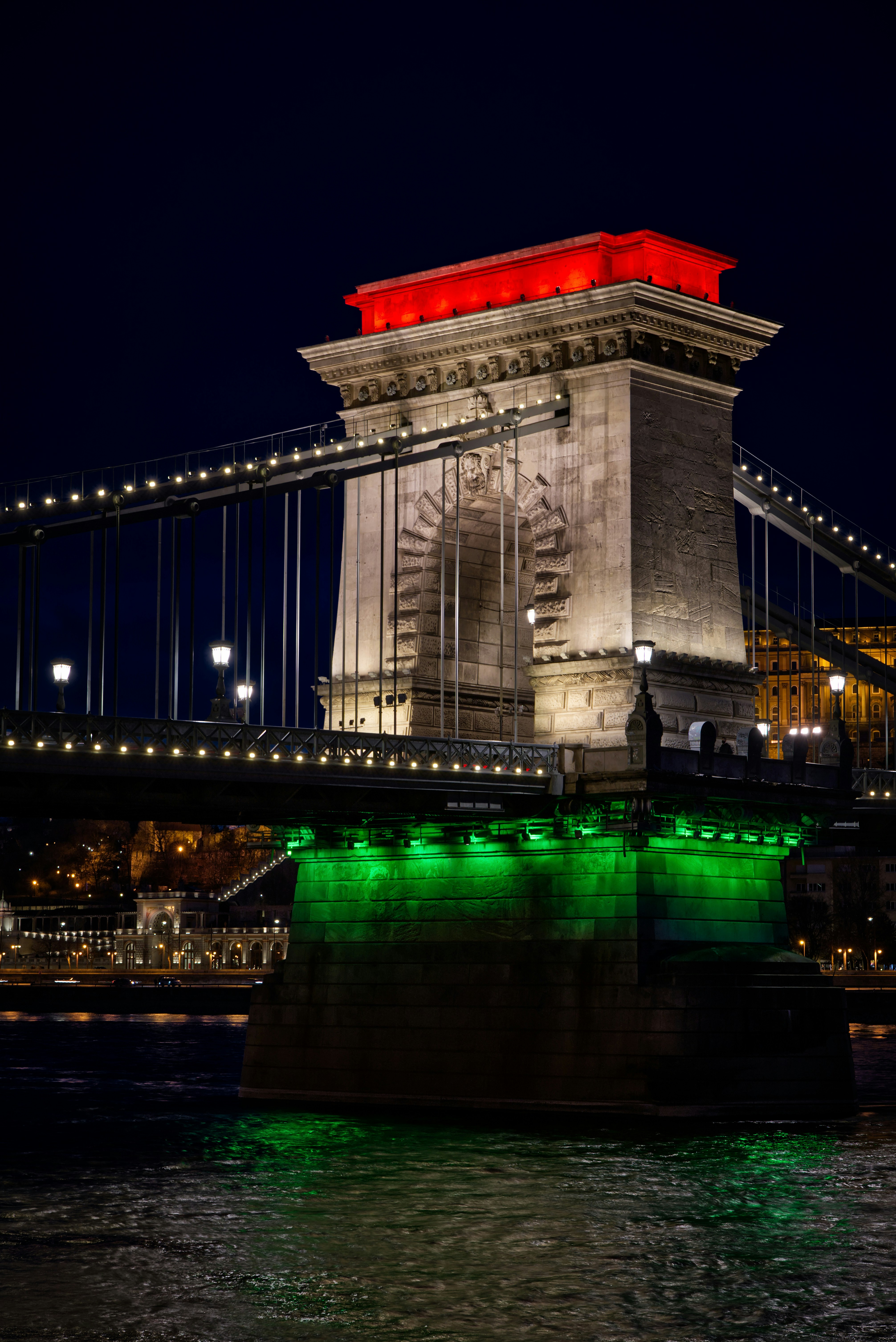 The Chain Bridge's iconic stone arch illuminated in festive colors against a dark sky, reflecting on the water below.