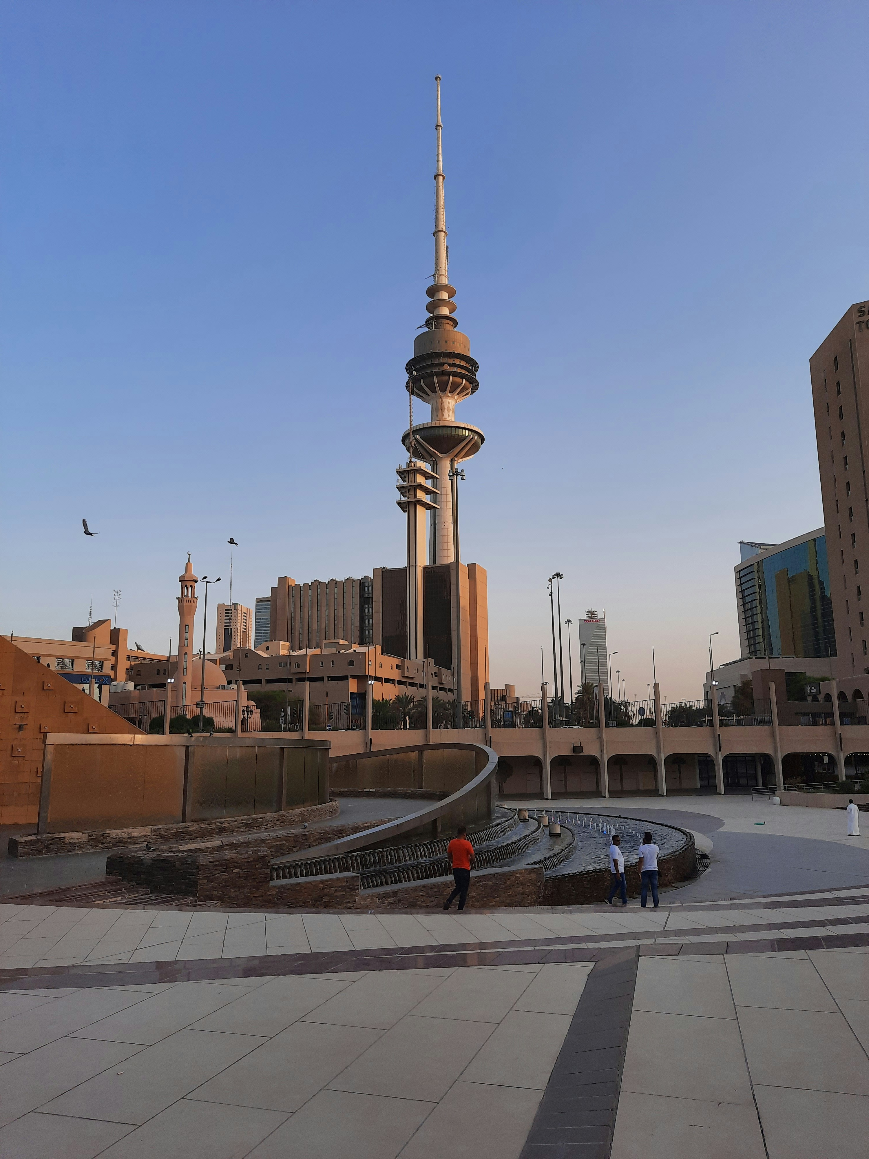 Kuwait's iconic telecommunications tower rises against a pastel sky, surrounded by contemporary architecture and people enjoying the plaza.