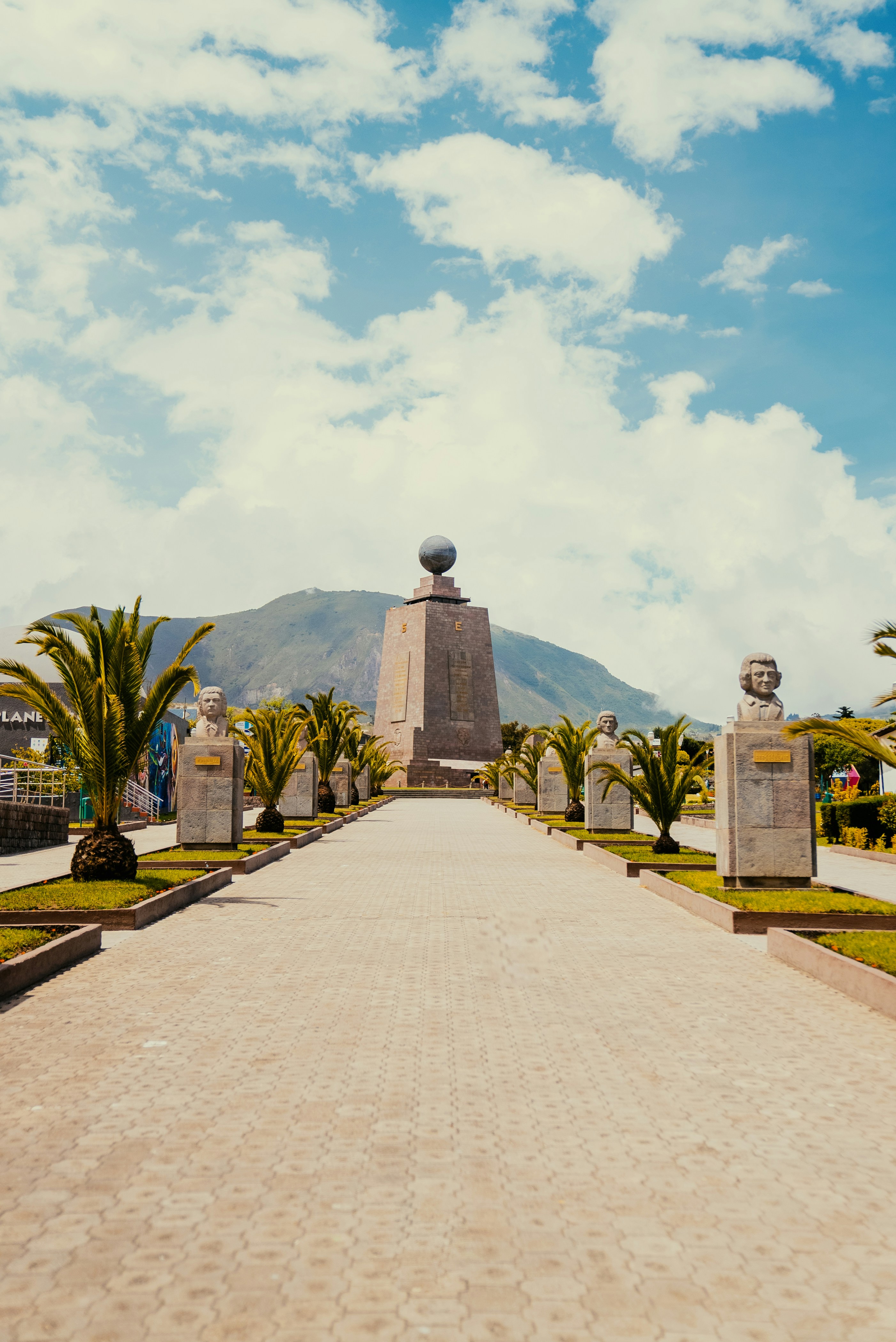Monumental obelisk surrounded by palm trees and busts, leading towards a distant mountain under a vibrant sky.